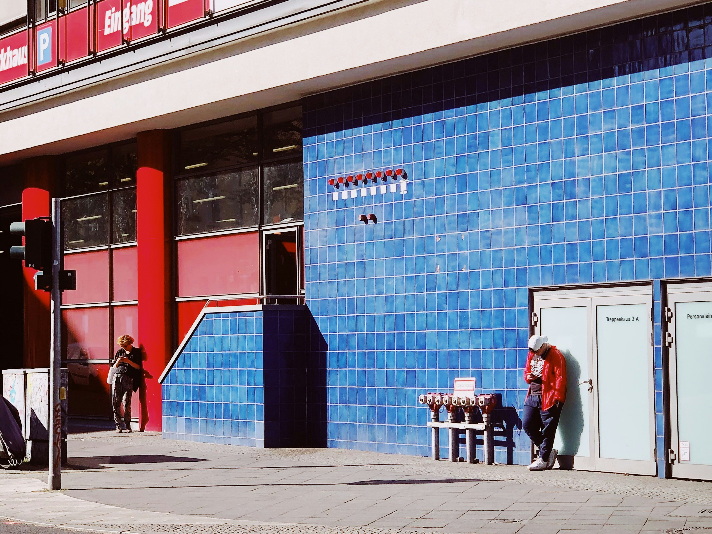 Vibrant blue tiled wall juxtaposed with red architectural elements and a lone figure leaning against a door. The scene captures a moment of urban life.