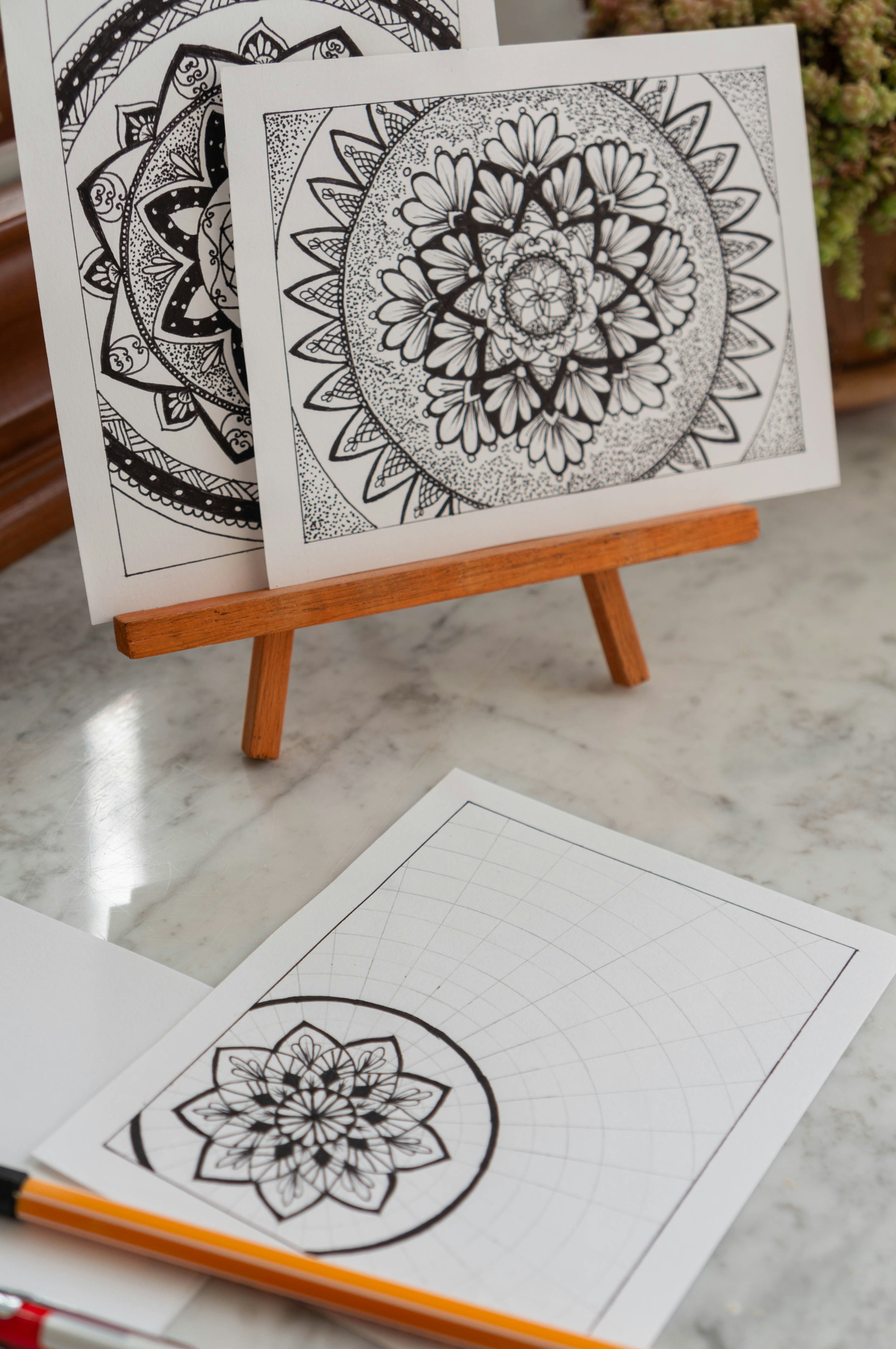 Two black-and-white mandala illustrations displayed on a small wooden easel, with a grid-lined sheet showing a partial mandala nearby on a marble table.