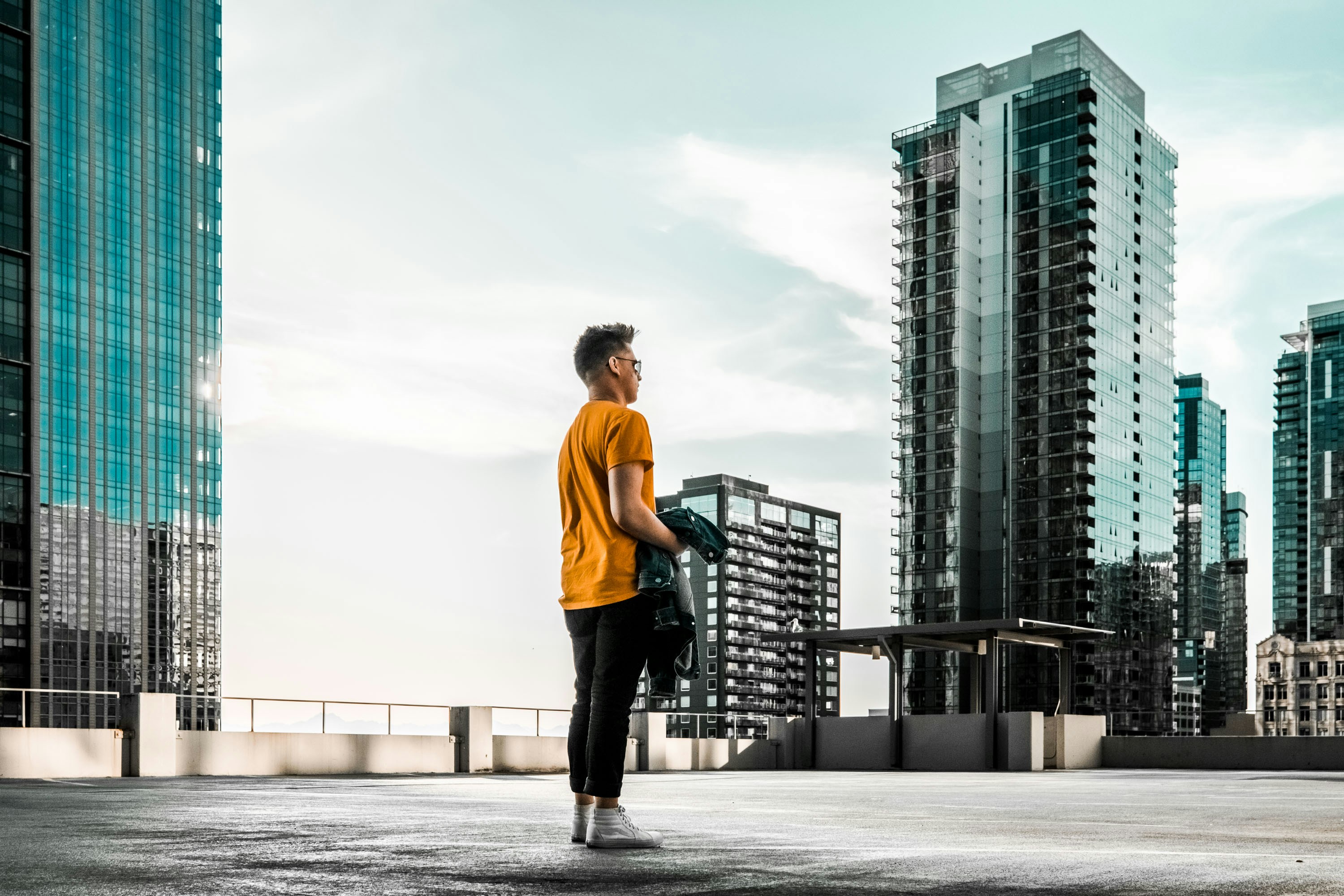 Individual in a bright orange shirt stands on a rooftop, gazing at towering skyscrapers under a clear sky.