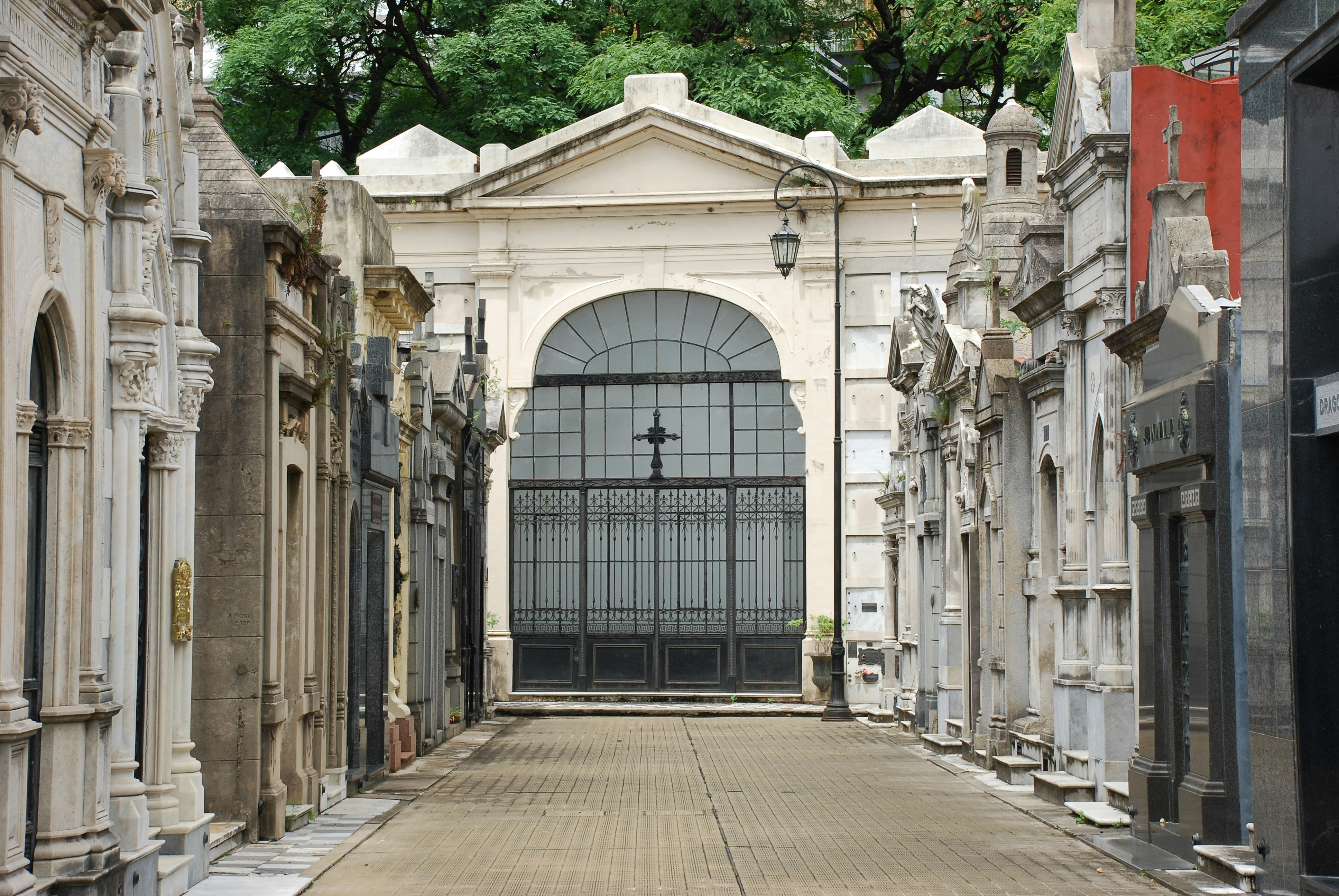 Tree-lined cemetery avenue flanked by ornate mausoleums under a cloudy sky.