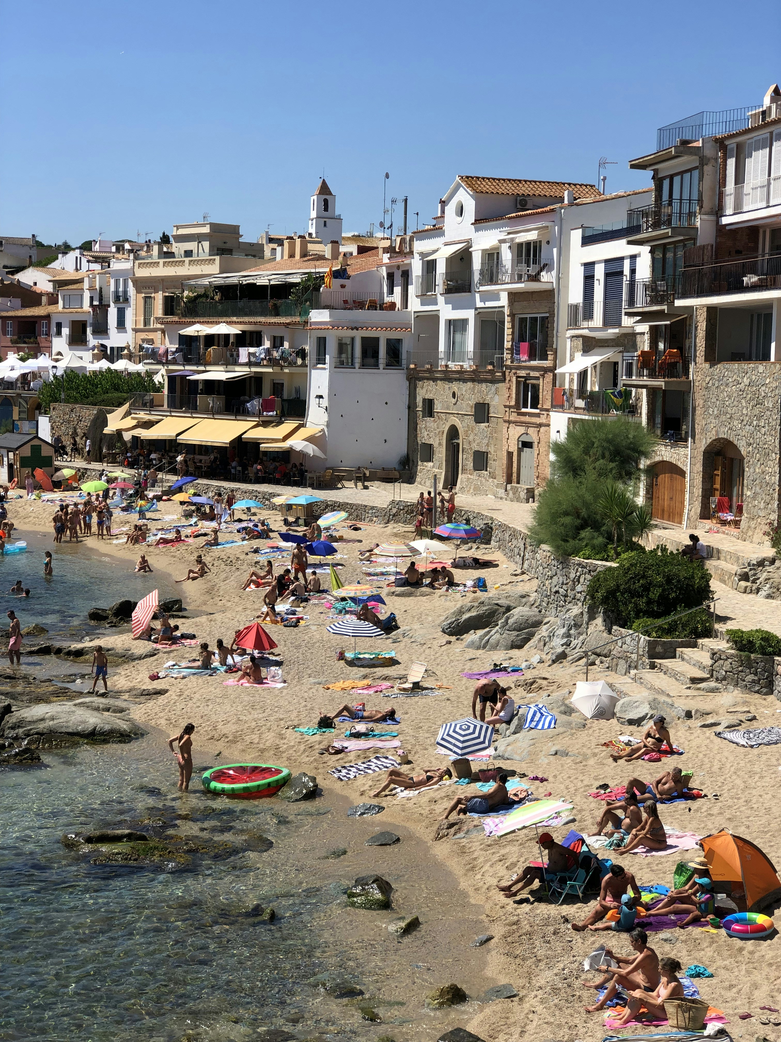 Sunlit Beachfront Odyssey: Crowded Pebble Shores Beneath Whitewashed Village Balconies