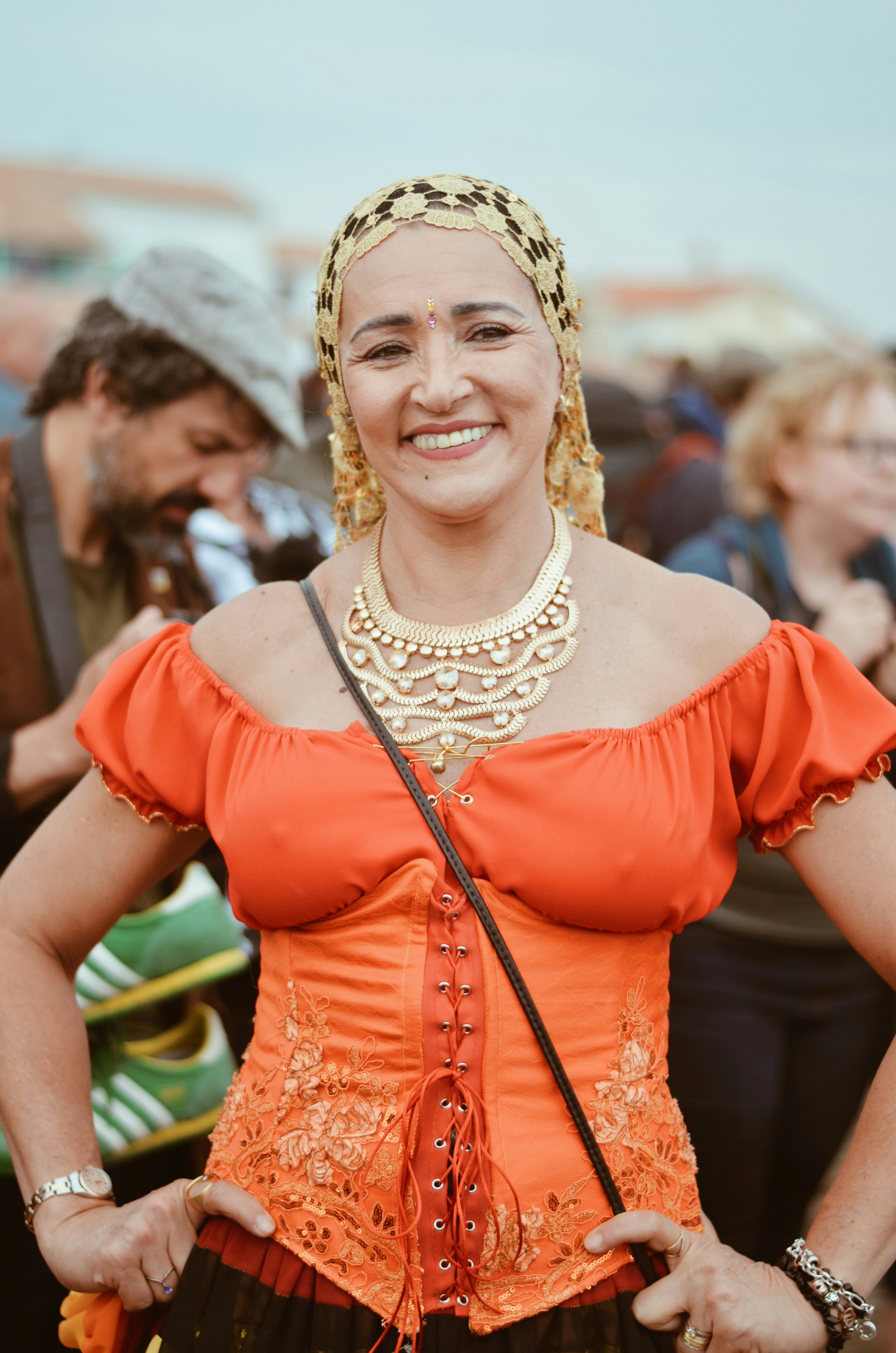 A smiling woman in a vibrant orange outfit adorned with intricate jewelry, exuding warmth and confidence amidst a lively crowd.