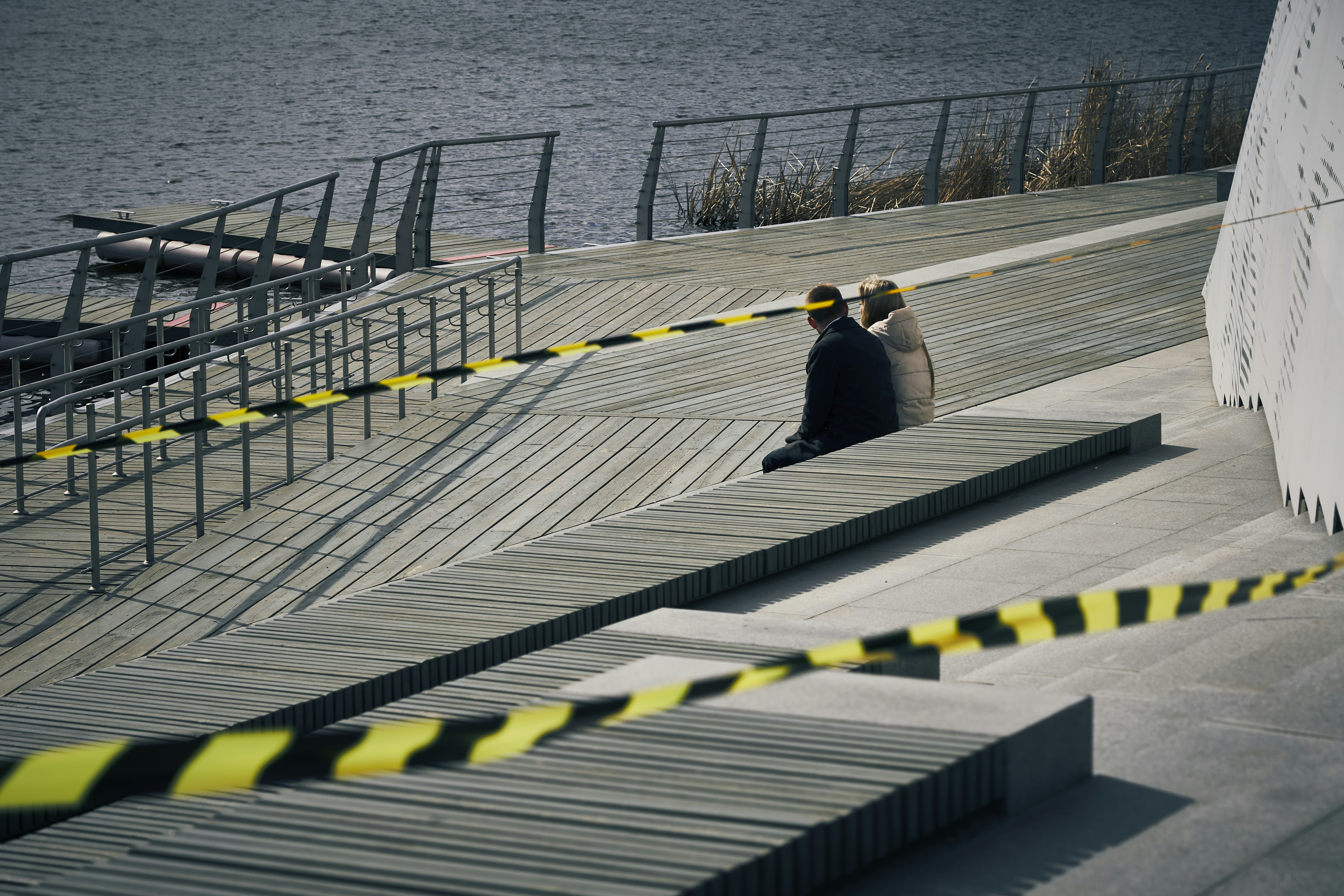 Quiet Harbor Pause: Two Figures Rest on Slatted Steps Under Caution Tape