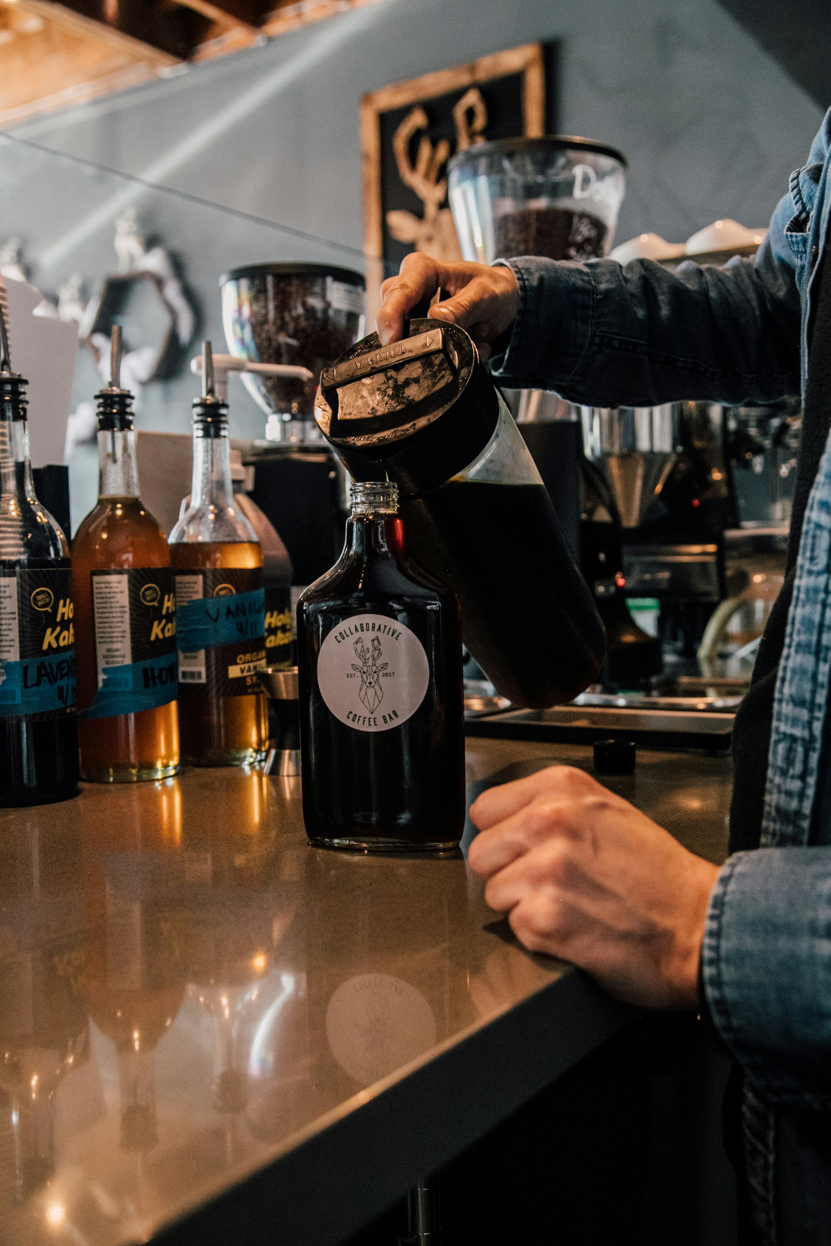 Barista pouring cold brew coffee from a French press into a glass bottle on a polished countertop, with various syrups in the background.