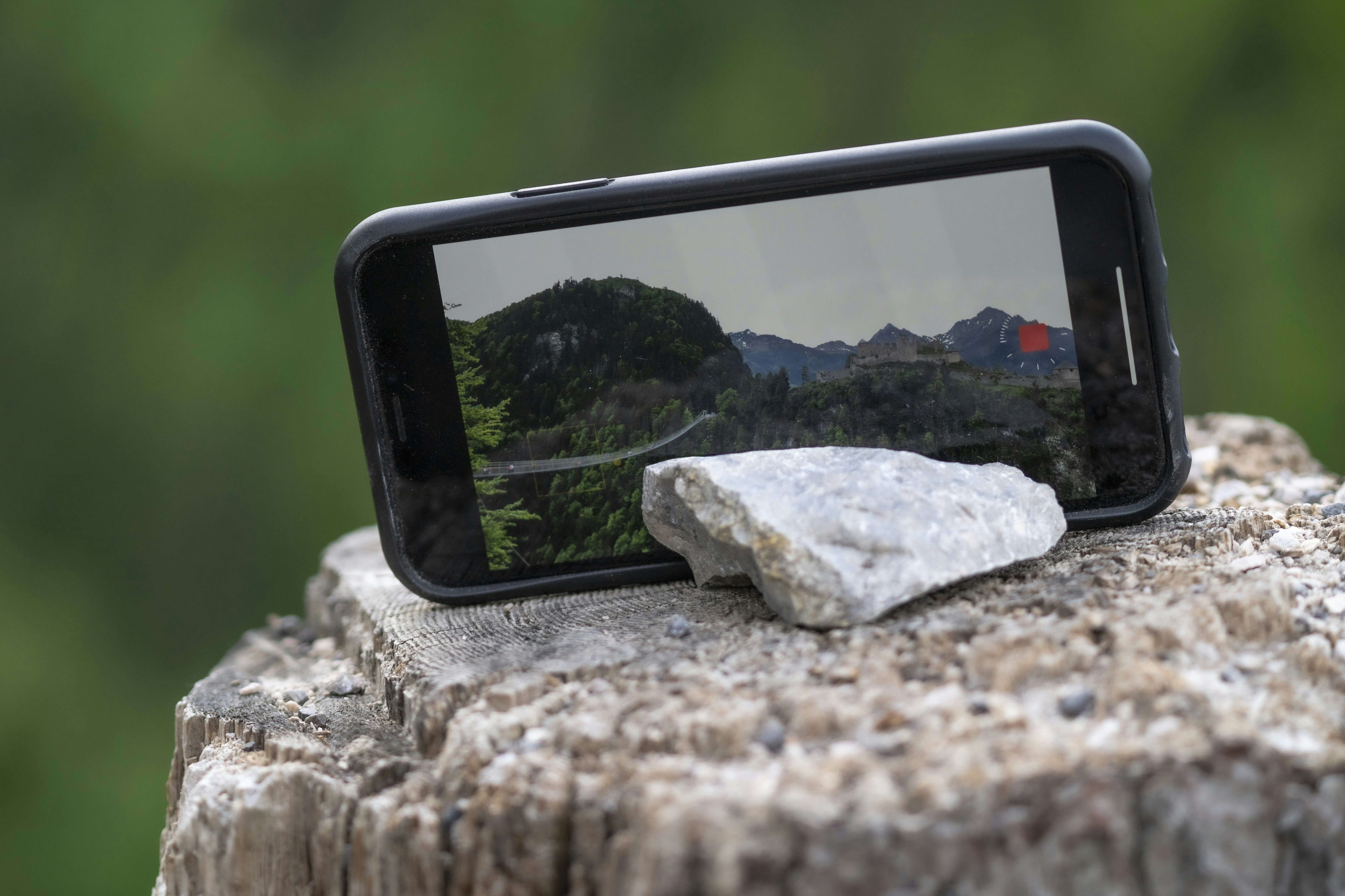 Smartphone resting on a jagged rock, screen shows a forested landscape with distant mountains. The shallow depth of field keeps the device in sharp focus against a blurred green backdrop.