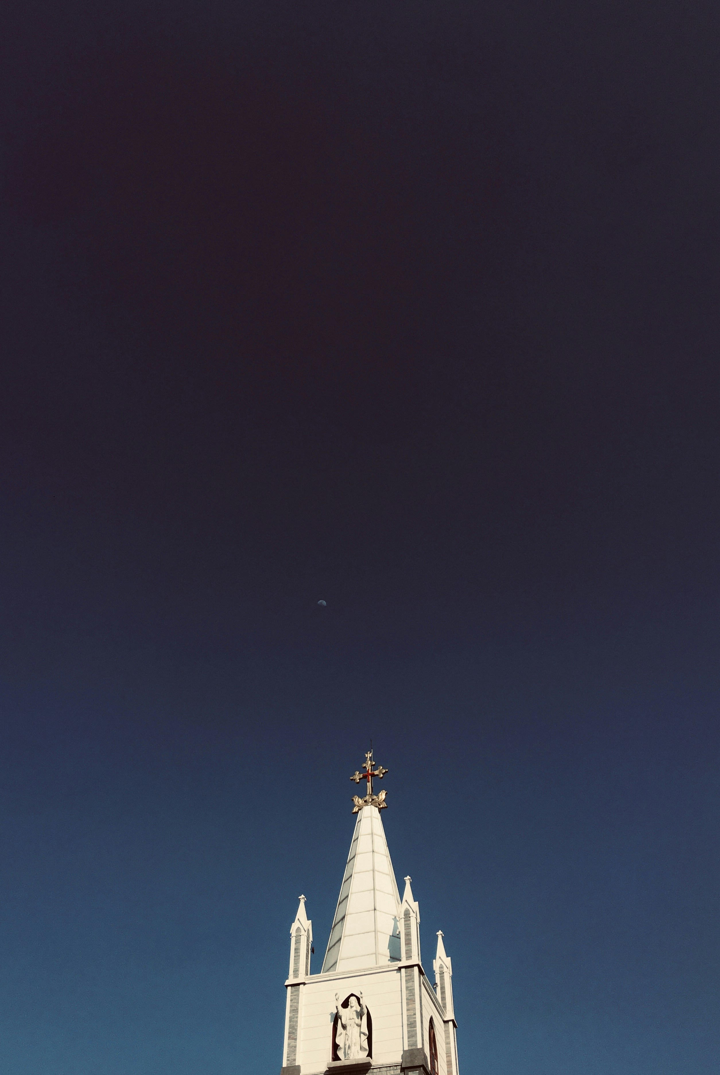White and yellow church steeple topped with a cross under a deep blue sky with a faint moon.