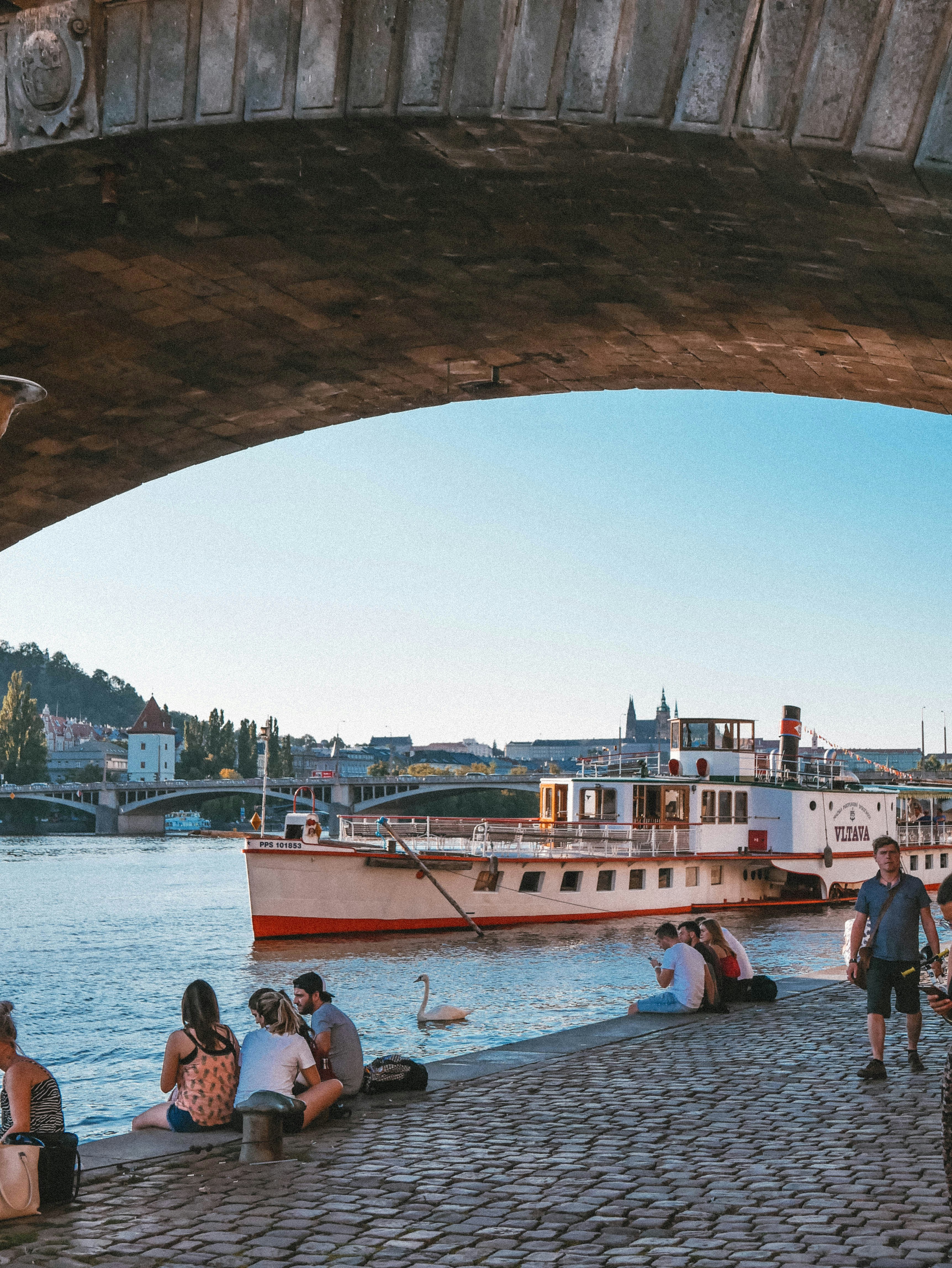 People relax along the riverbank under a stone archway, while a vintage boat floats gracefully on the water. A swan glides nearby.