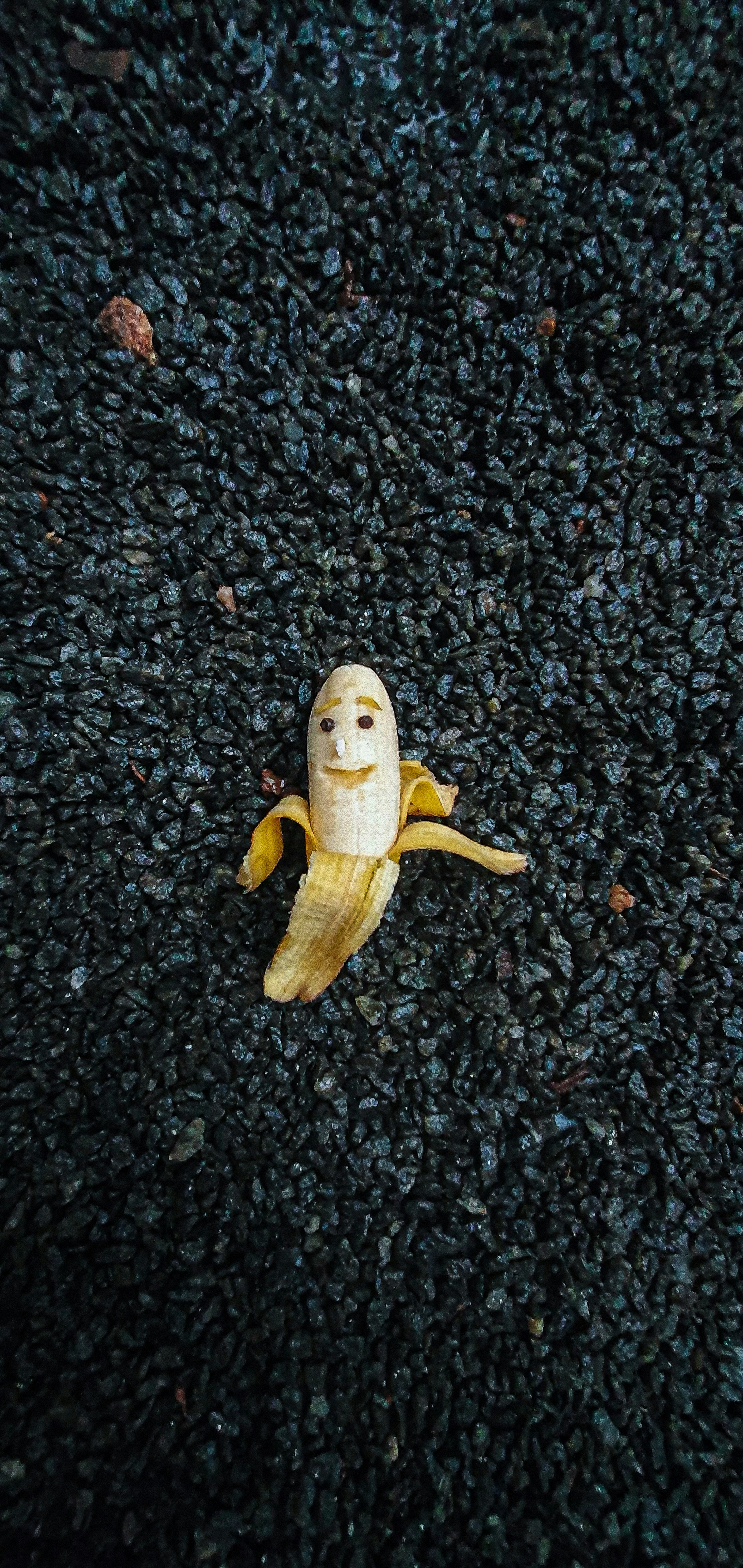 Close-up photograph of a peeled banana with a simple face drawn on it, resting on dark, pebbly asphalt.
