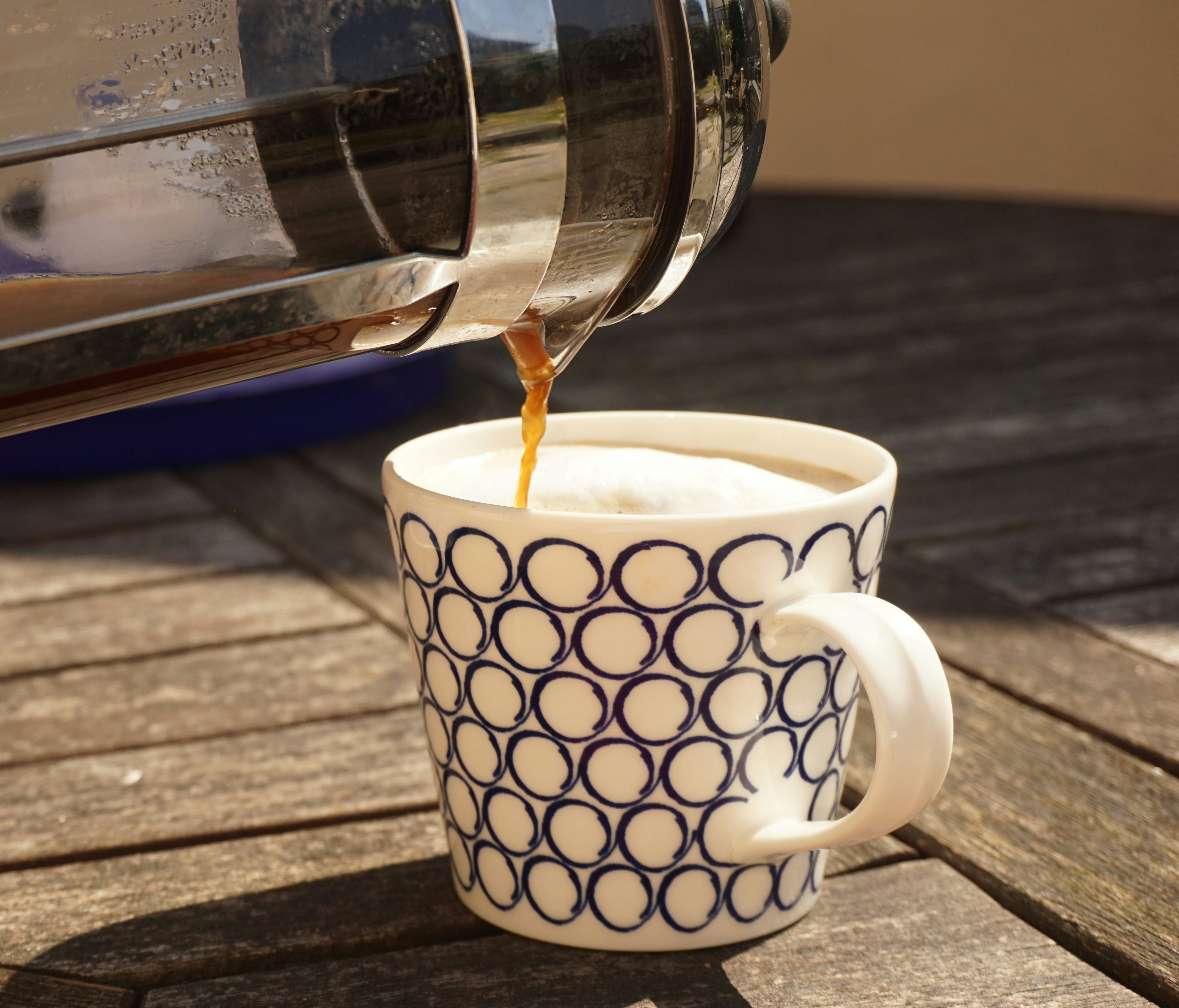 Freshly brewed coffee being poured from a French press into a patterned mug on a wooden table.