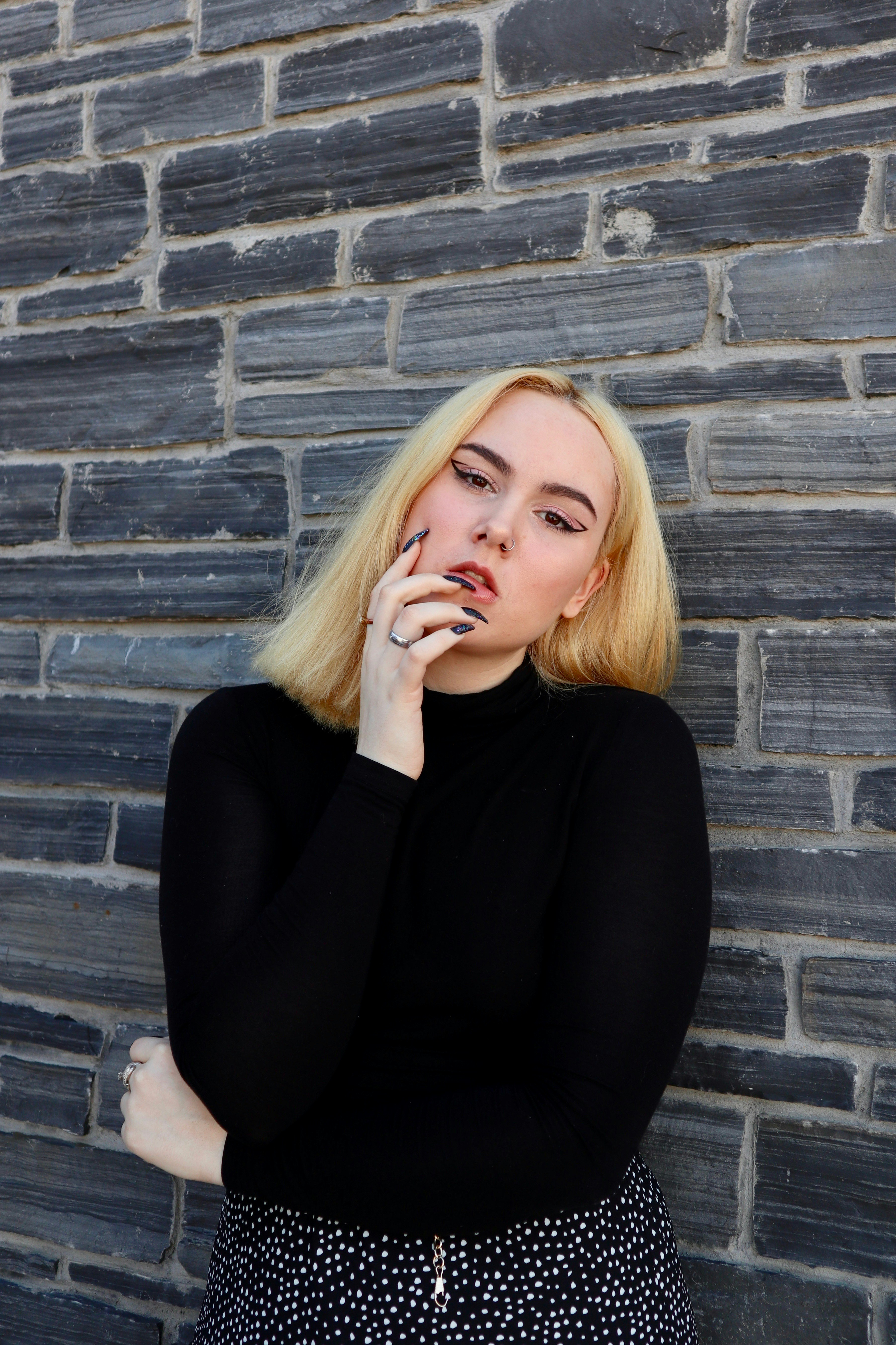 Portrait photograph of a blonde woman in a black turtleneck posed against a textured gray brick wall. This shot emphasizes moody fashion and urban atmosphere.