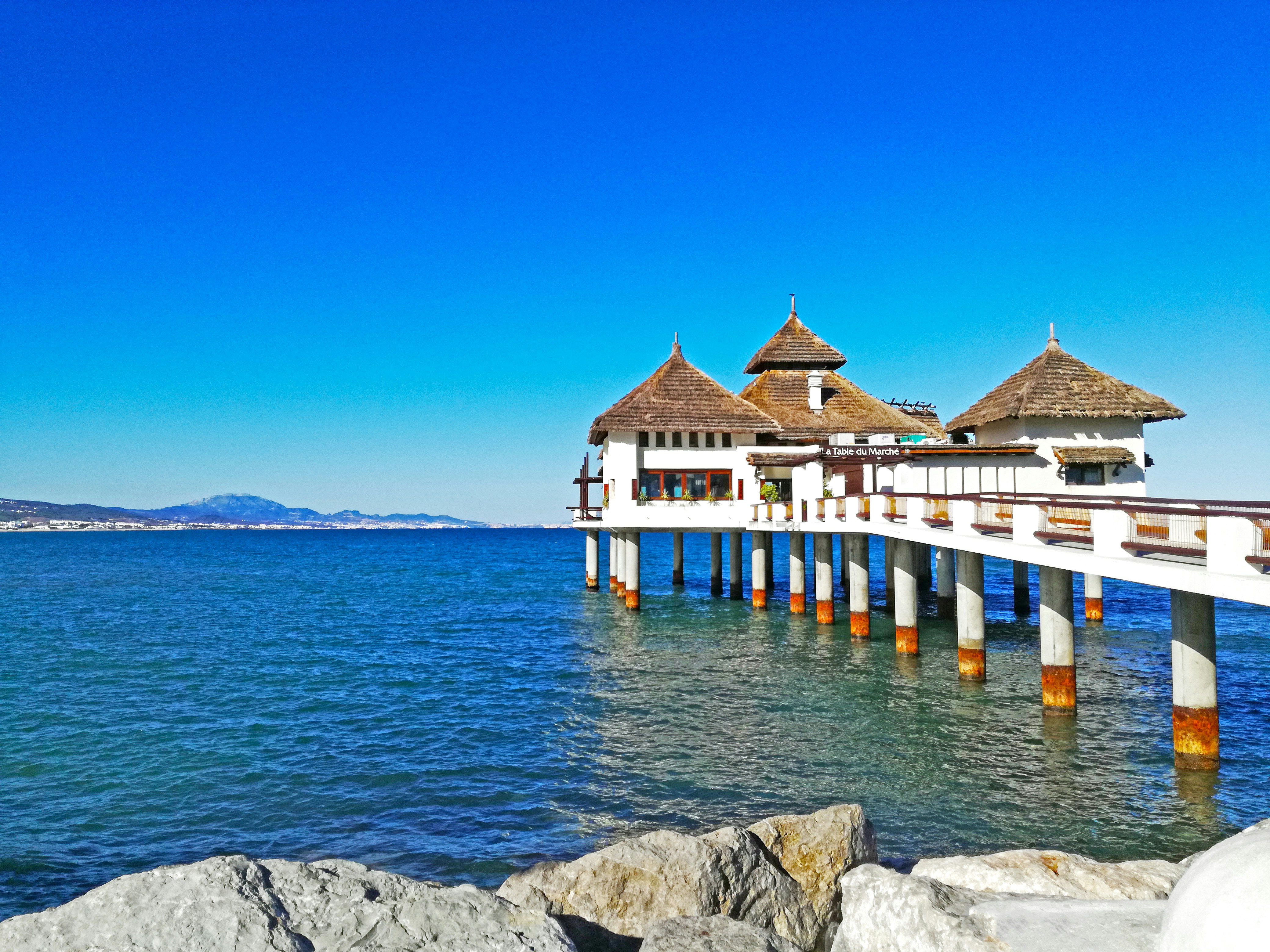Pier with thatched-roof structure extends over clear blue water under a vibrant sky.