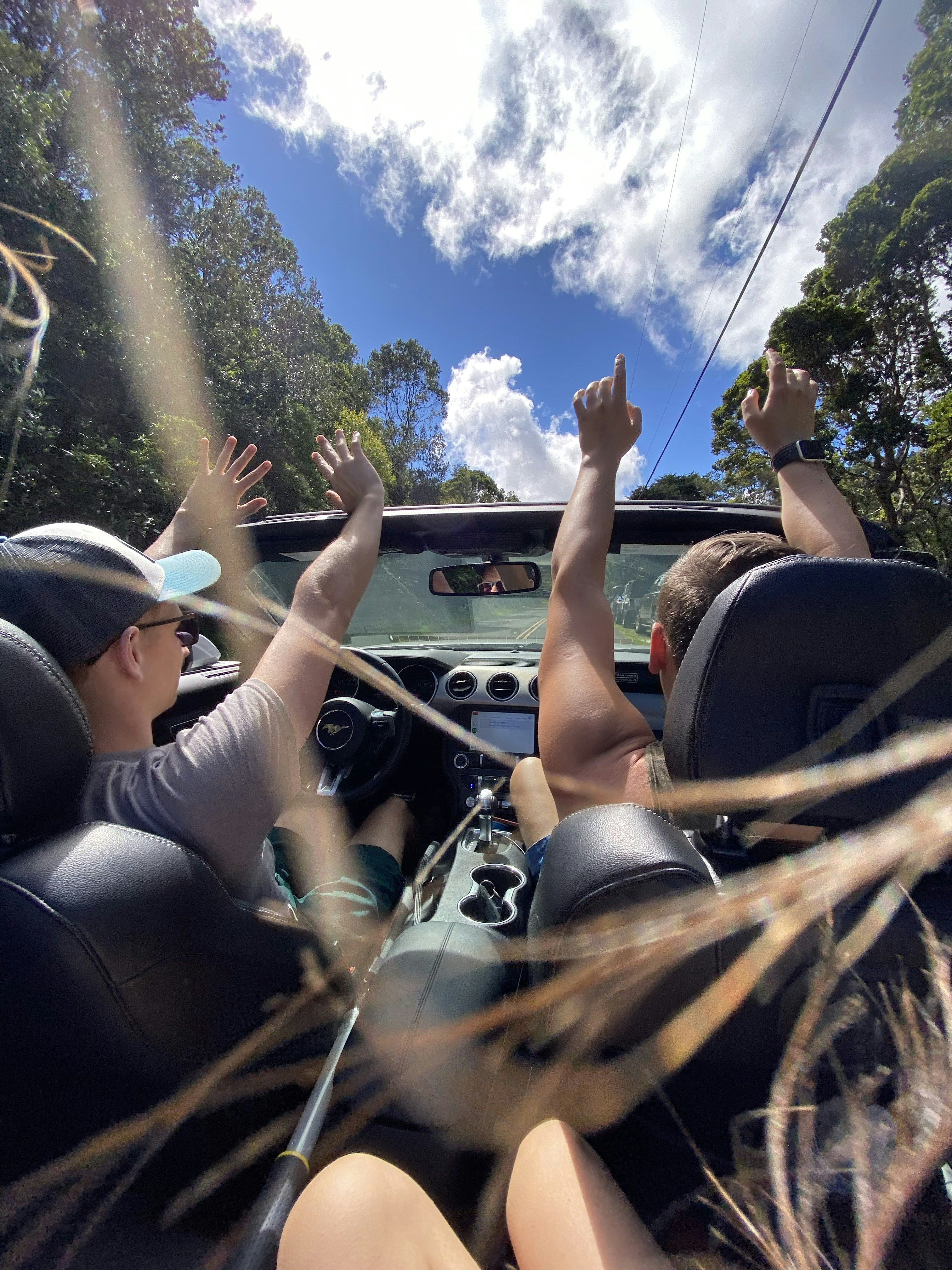 Friends celebrating a carefree moment in a convertible with arms raised, surrounded by lush greenery and a bright blue sky.