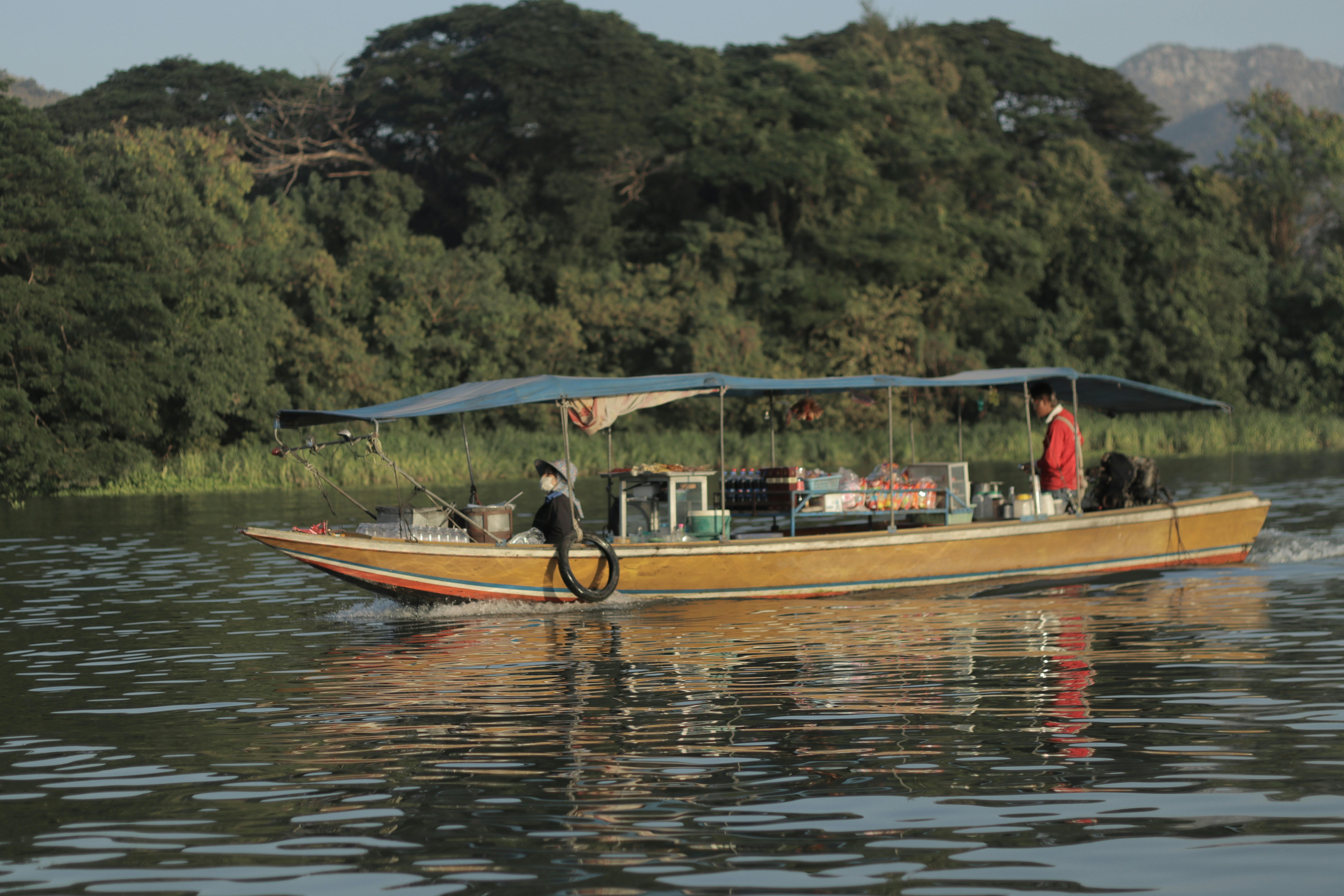A wooden boat glides across a serene lake, with vendors showcasing their goods under a shaded canopy. The reflection in the water adds a tranquil touch.