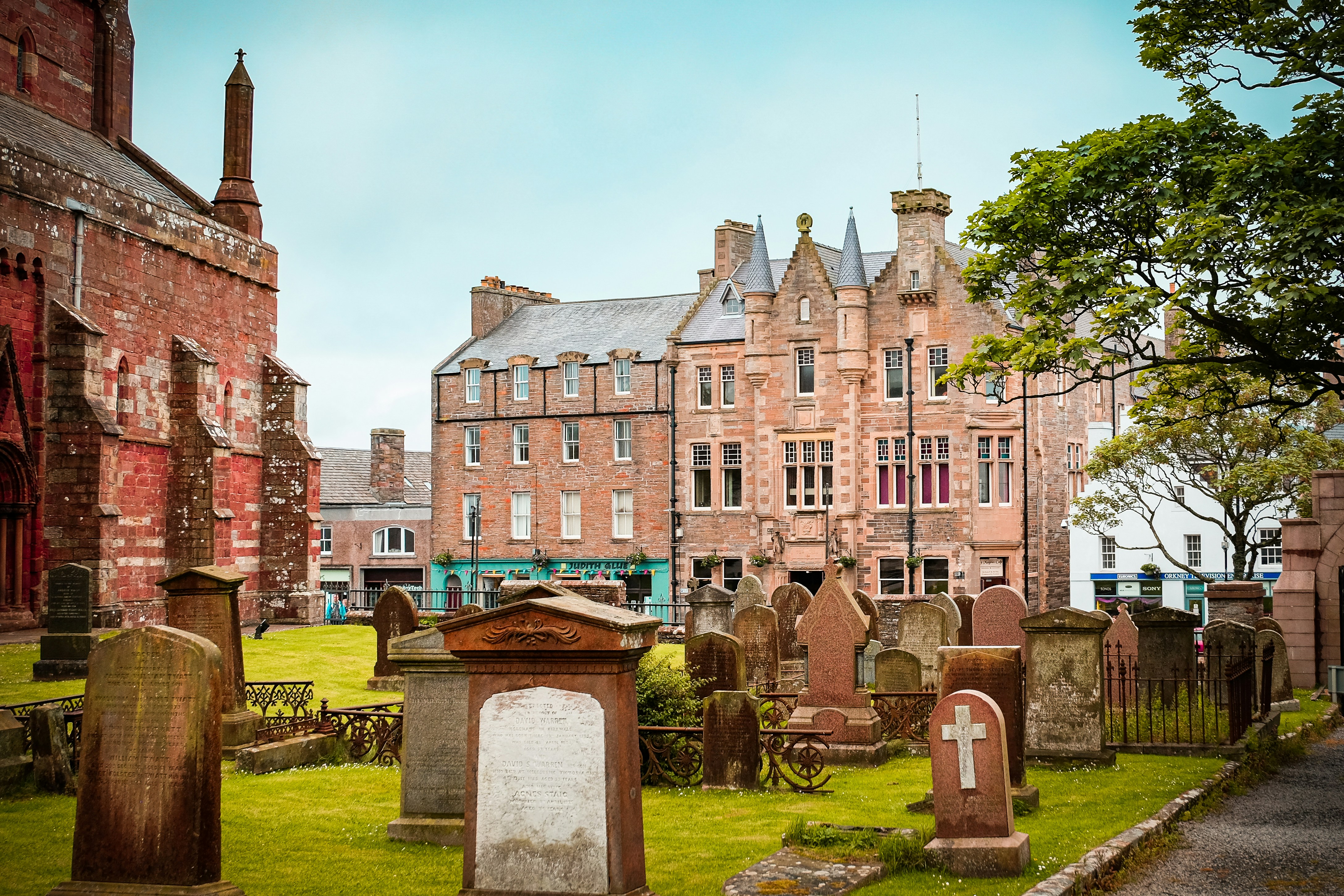 Ancient gravestones in a green cemetery surrounded by historic brick buildings and a tree under a clear blue sky.