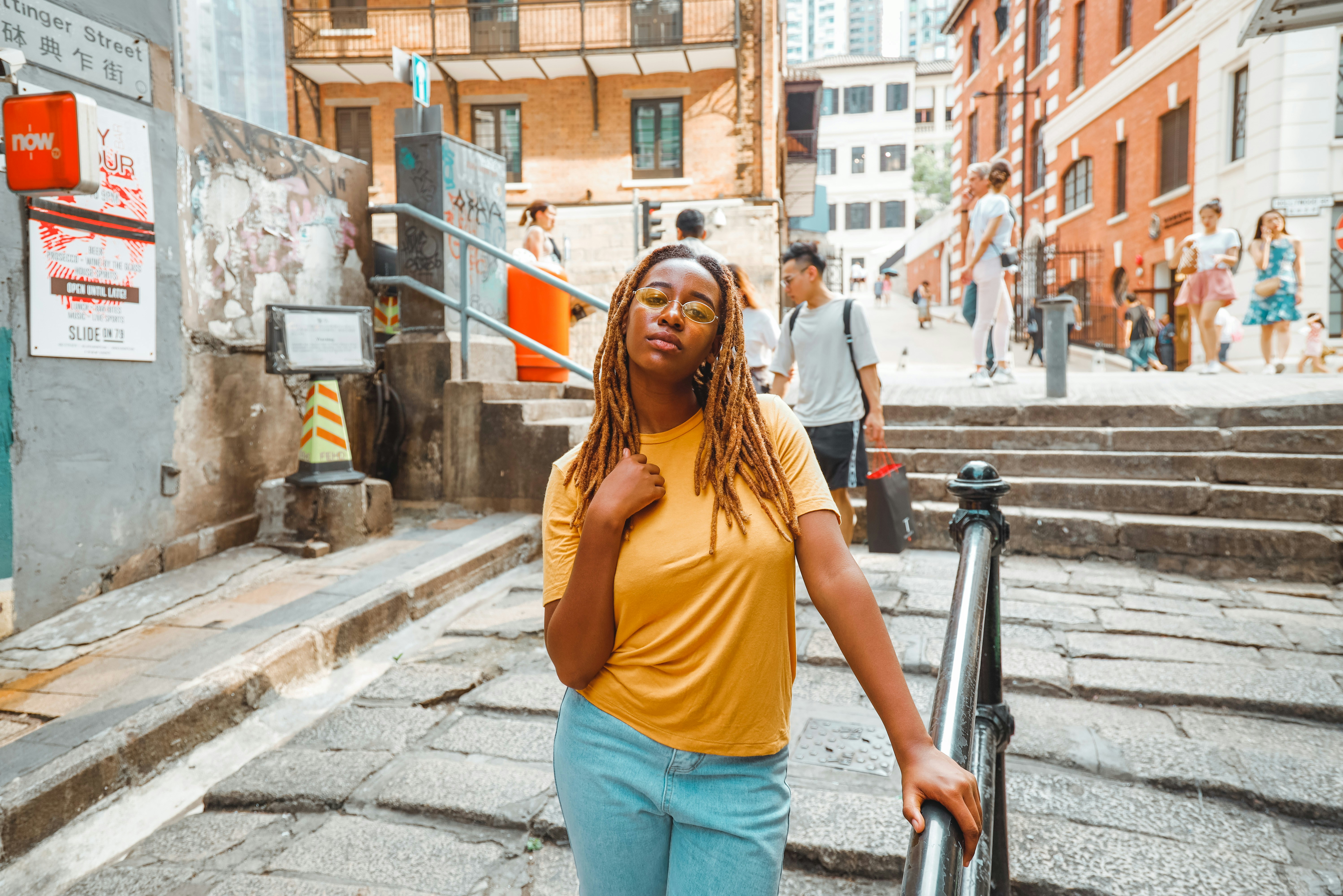 Person in orange shirt and blue jeans standing on a cobblestone street in a bustling urban setting.