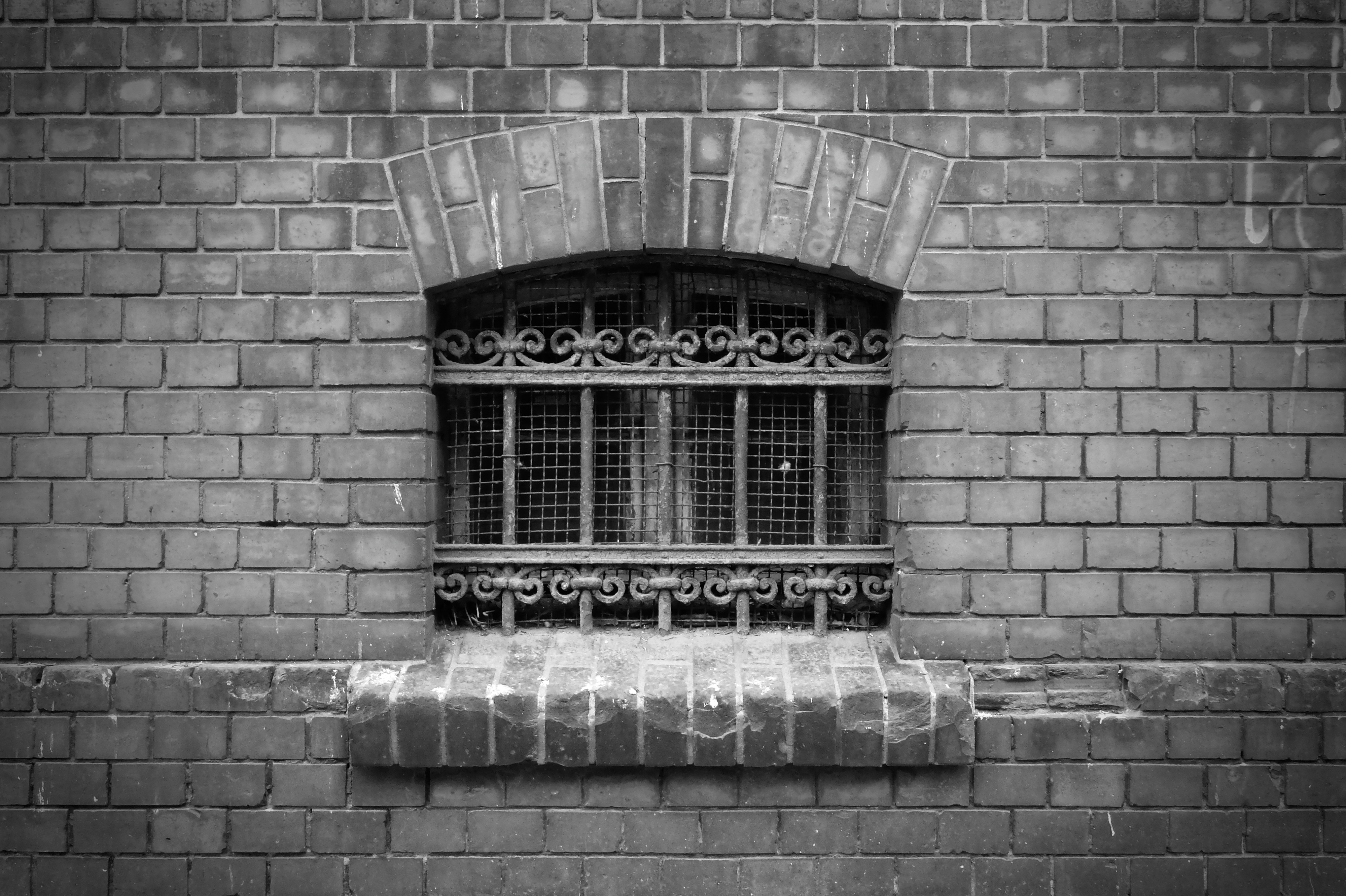 Grayscale view of an ornate iron-barred window set in a weathered brick wall.
