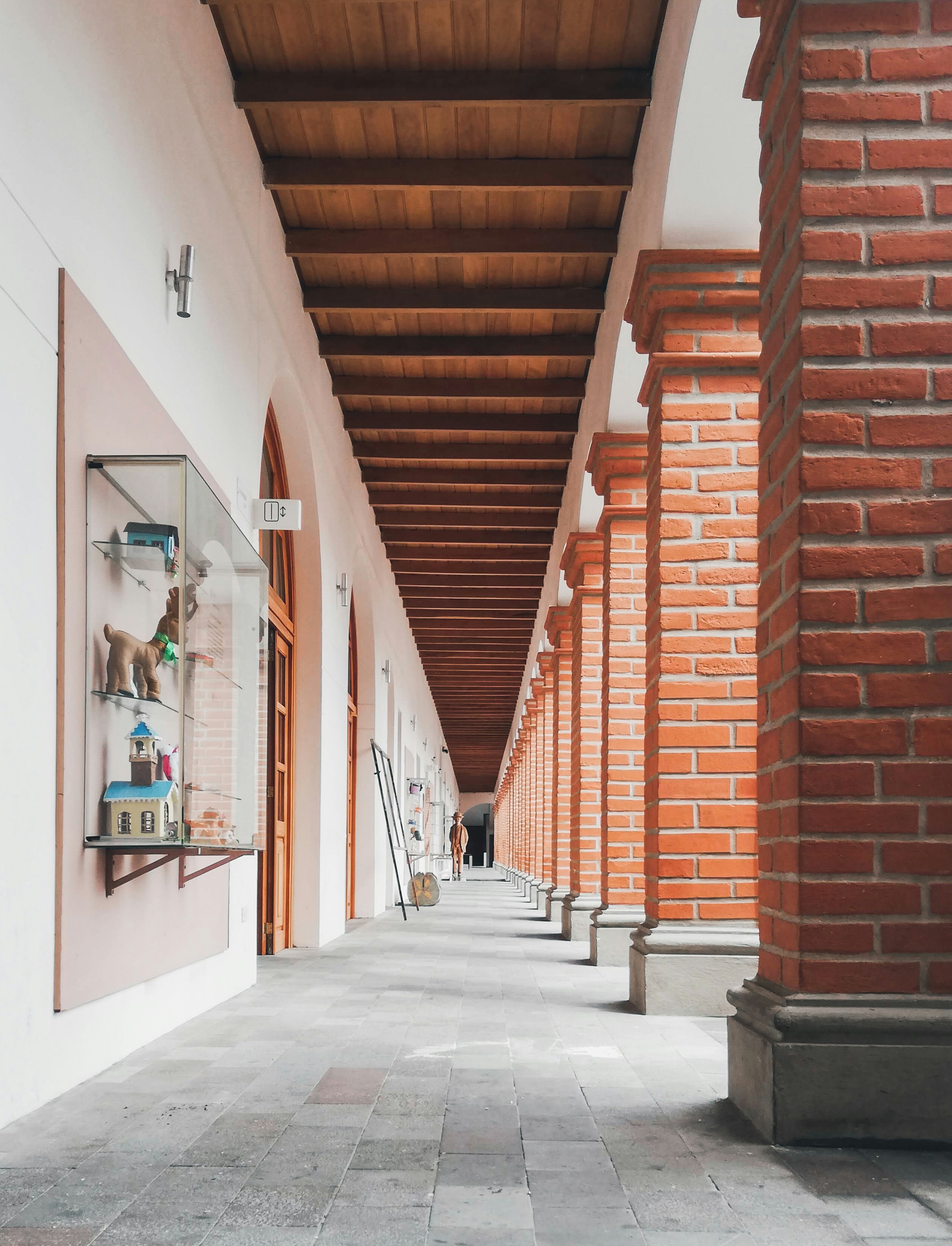 Long covered walkway with brick columns and a wooden beam ceiling receding toward a distant figure.