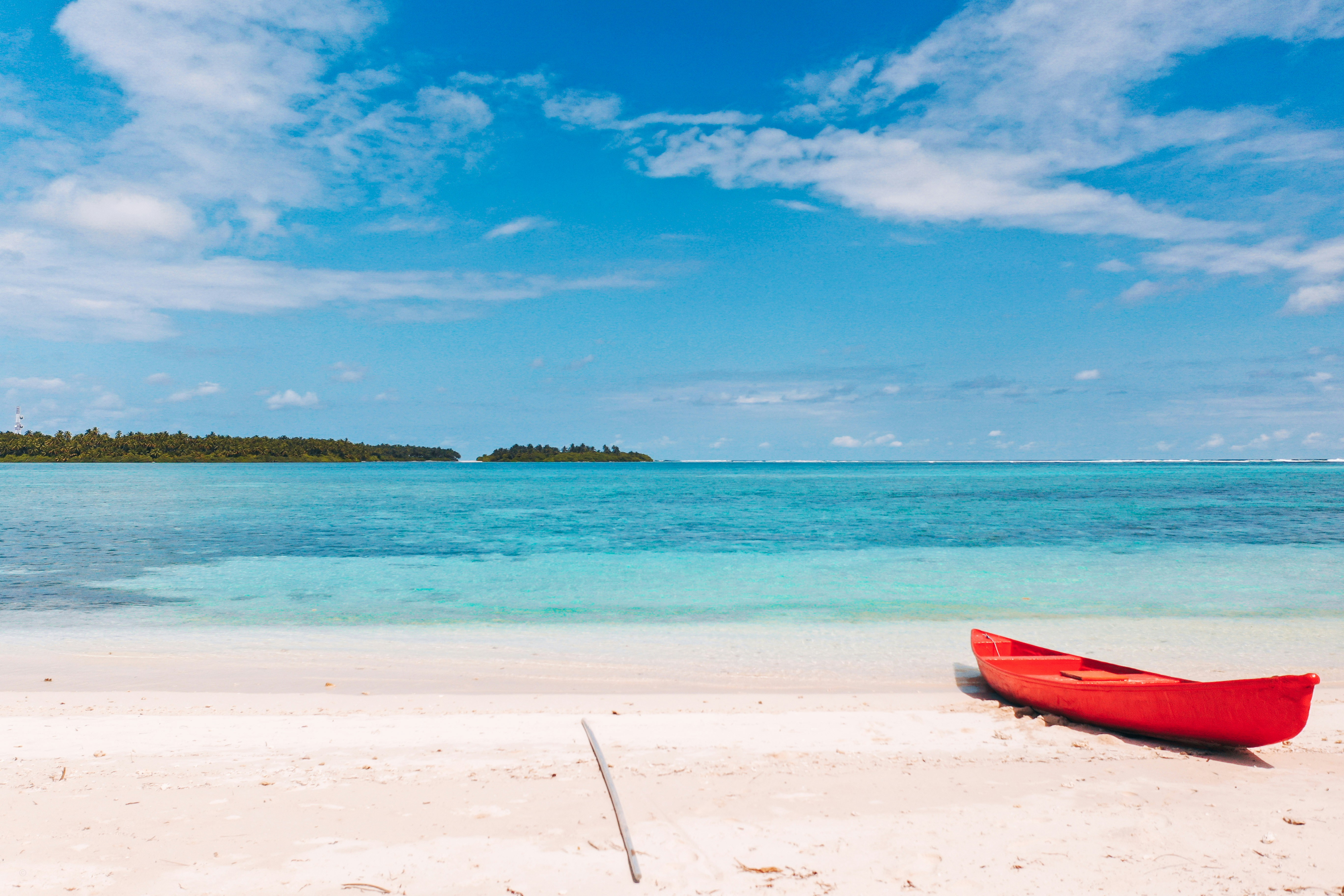 Red kayak resting on a pristine white sand beach with turquoise waters and distant islands under a bright blue sky.