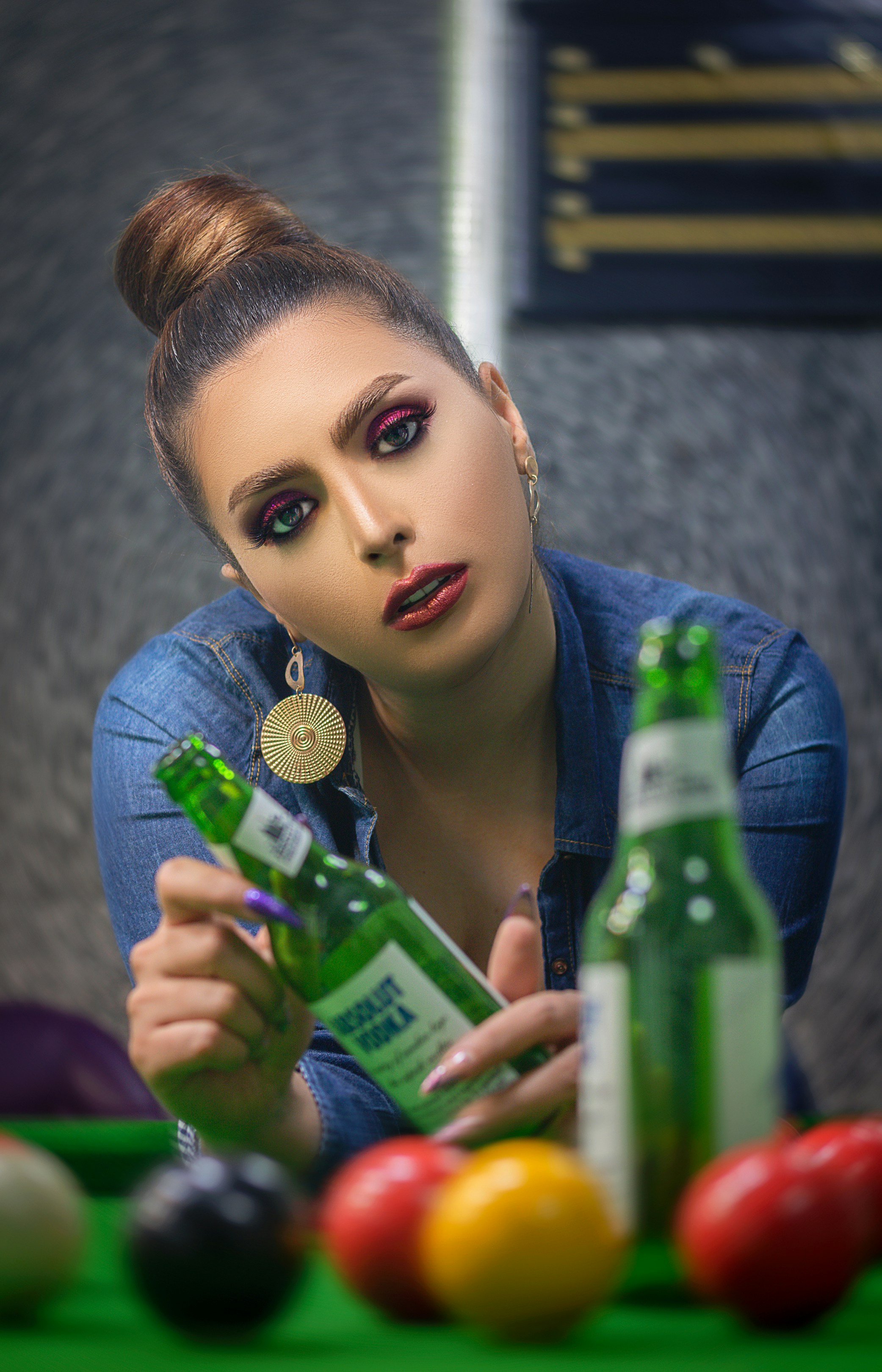 A woman with striking makeup holds a green bottle near a billiard table, surrounded by colorful balls. Her intense gaze draws attention to the scene's vibrant atmosphere.