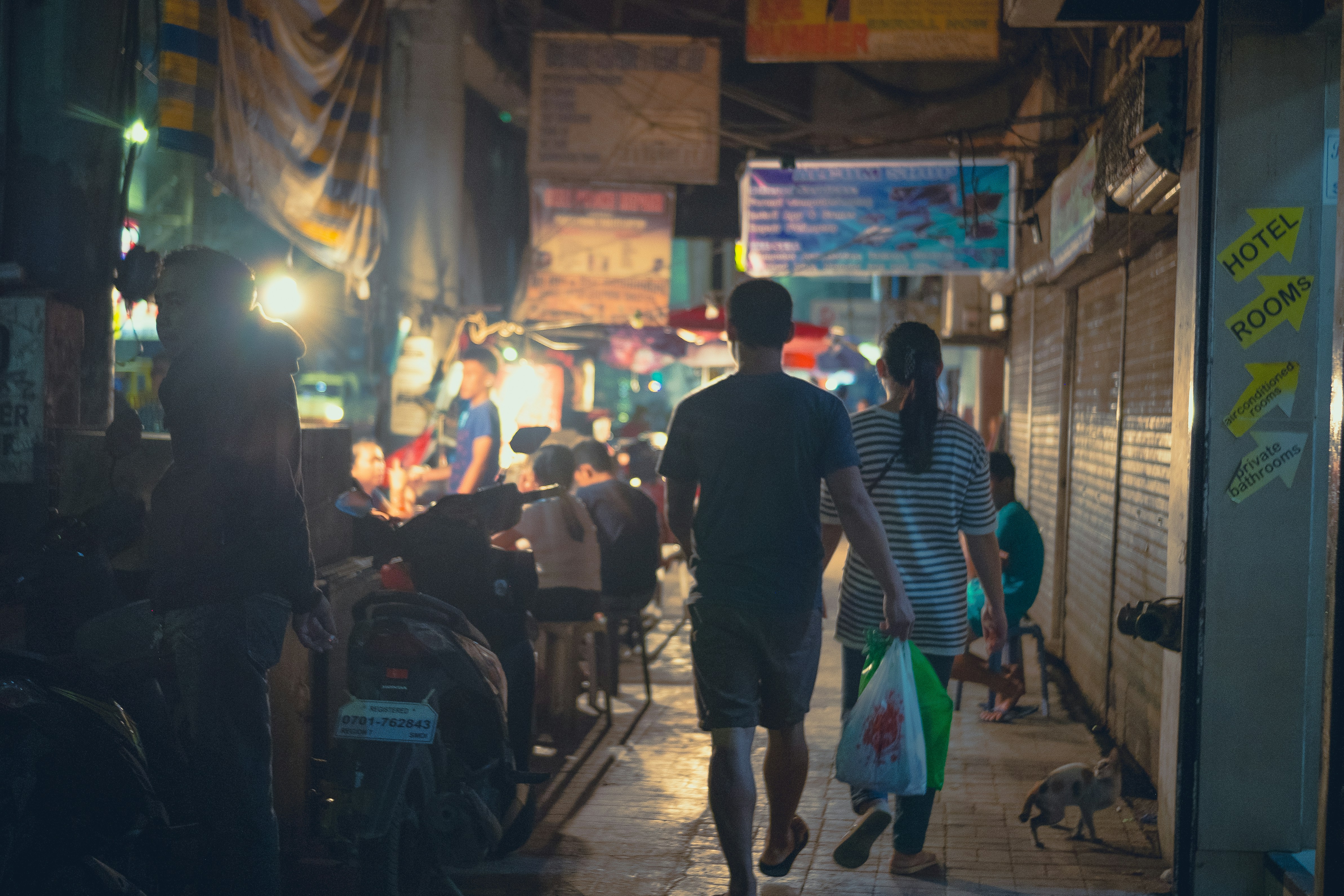 Couple walking through a bustling, dimly lit market alley with colorful signs and glowing lights.
