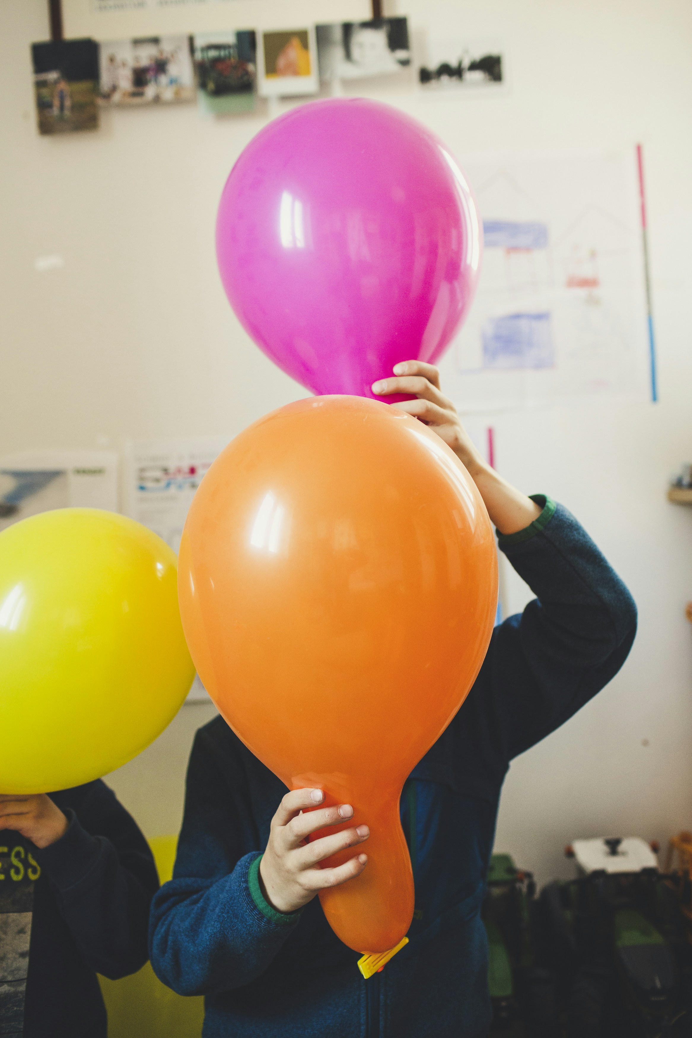 Children playfully hide behind vibrant red, yellow, and orange balloons indoors.