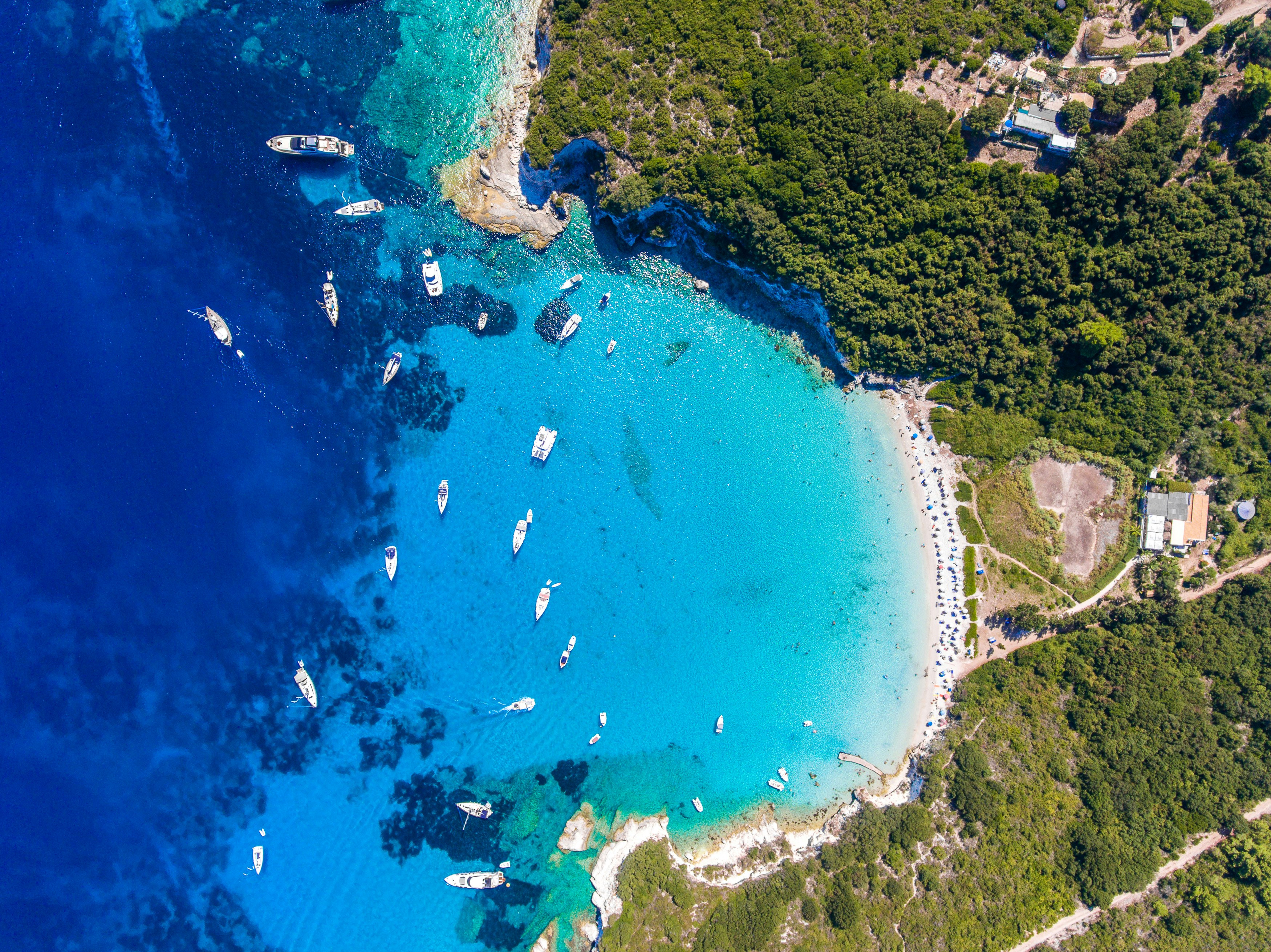 Turquoise waters of Antipaxos Island with yachts and a sandy beach surrounded by lush greenery.