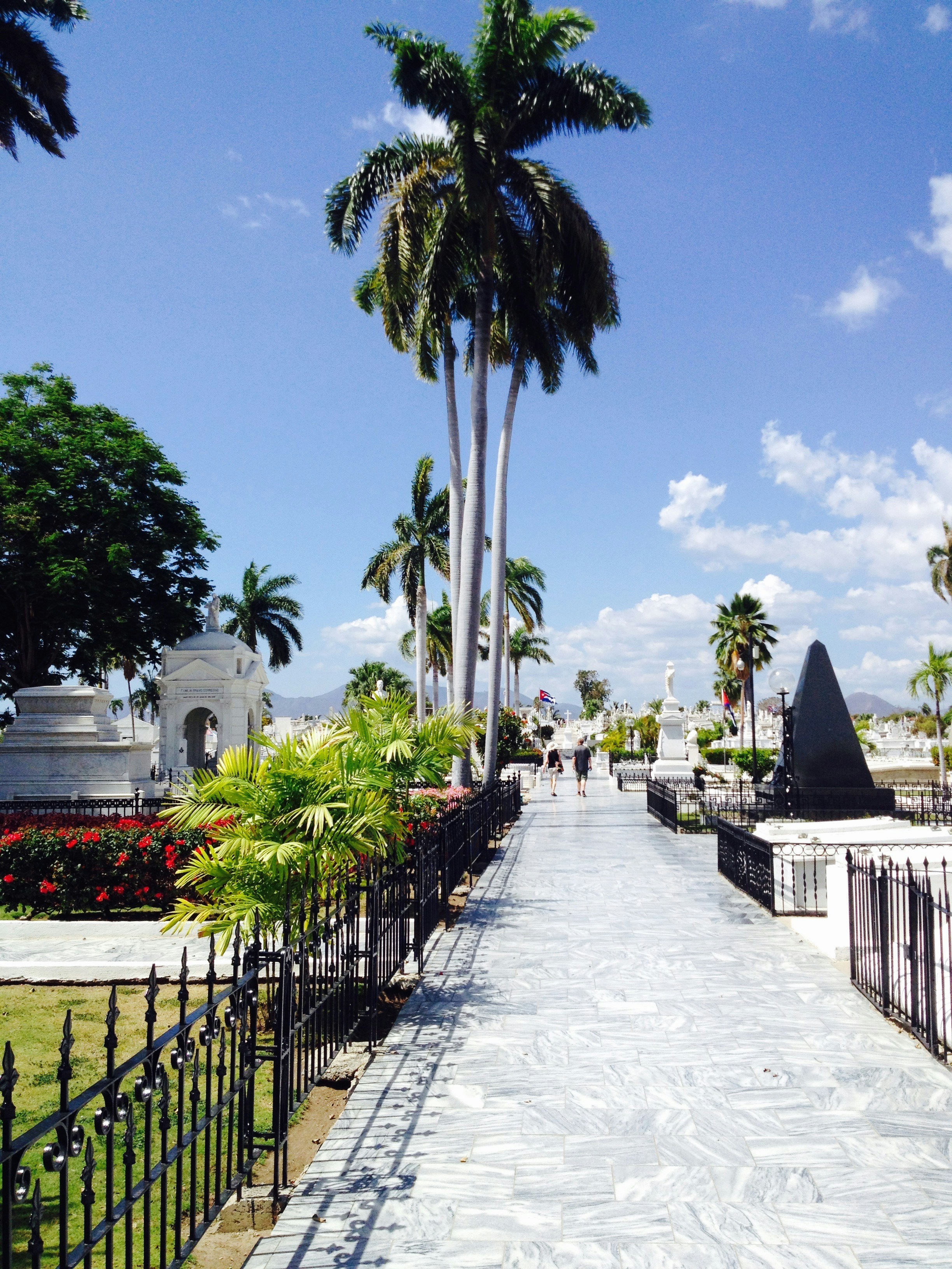 Lush palm trees line a marble pathway in a memorial garden, flanked by vibrant flowers and statues under a clear blue sky.