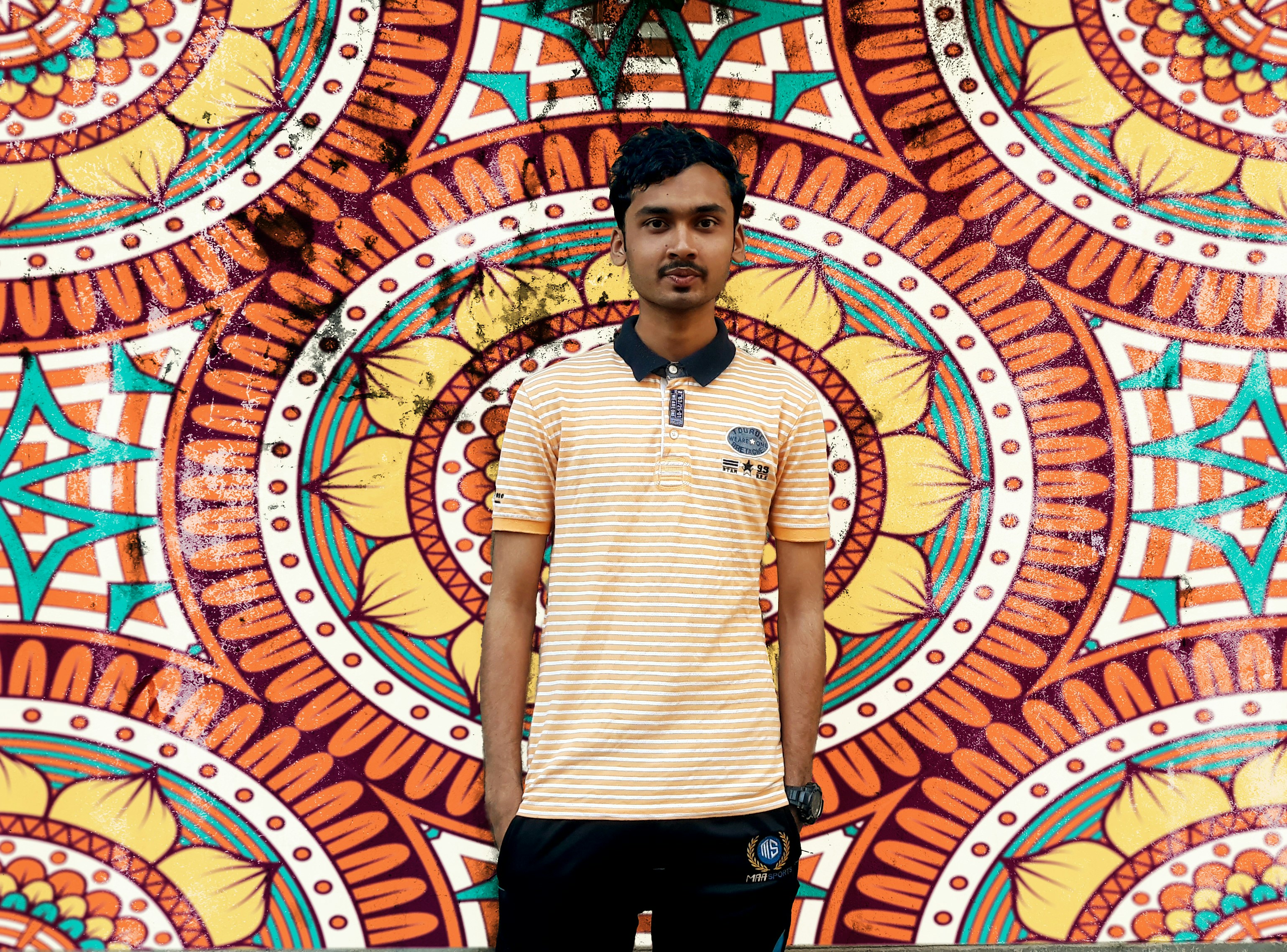 Man in a beige polo shirt standing against a colorful mandala mural with intricate patterns.