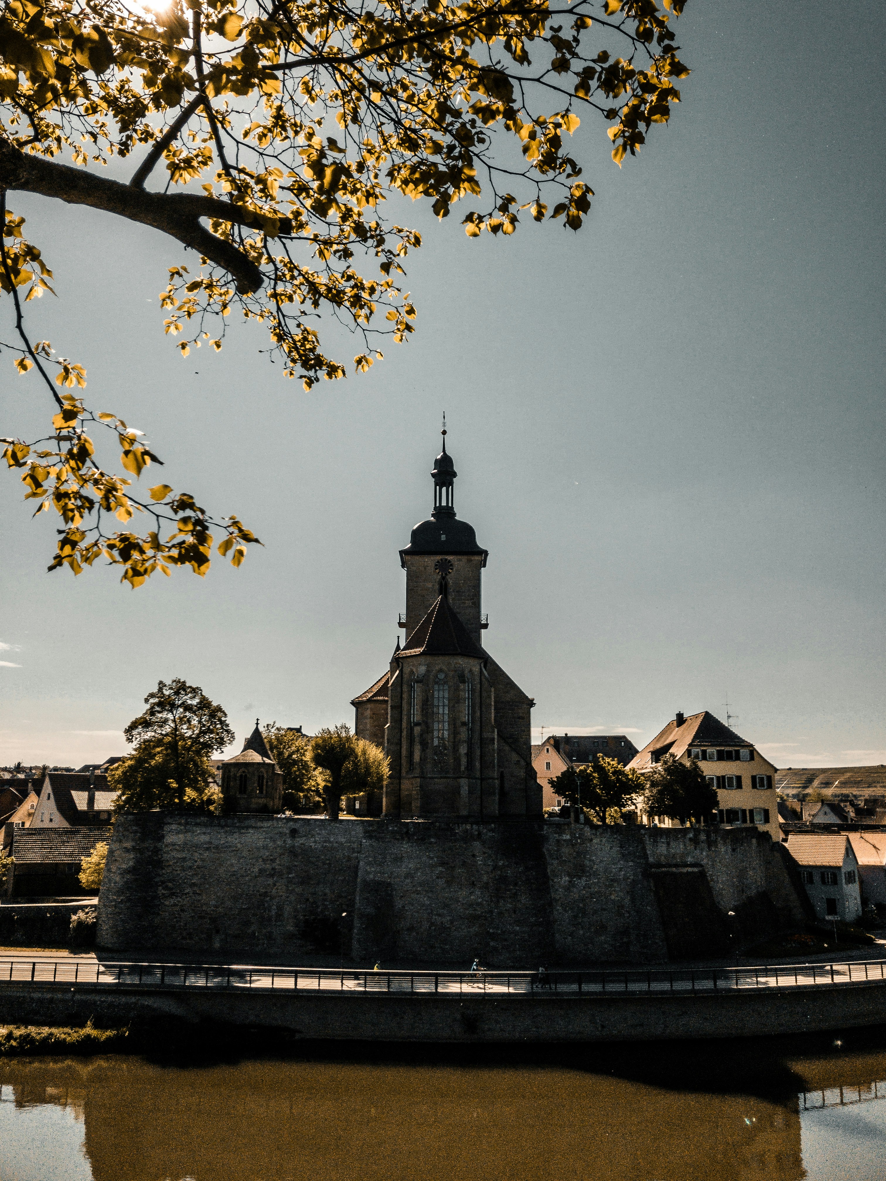 Historic church tower rises above a tranquil river, surrounded by lush trees and quaint buildings. The serene landscape reflects a blend of nature and architecture.
