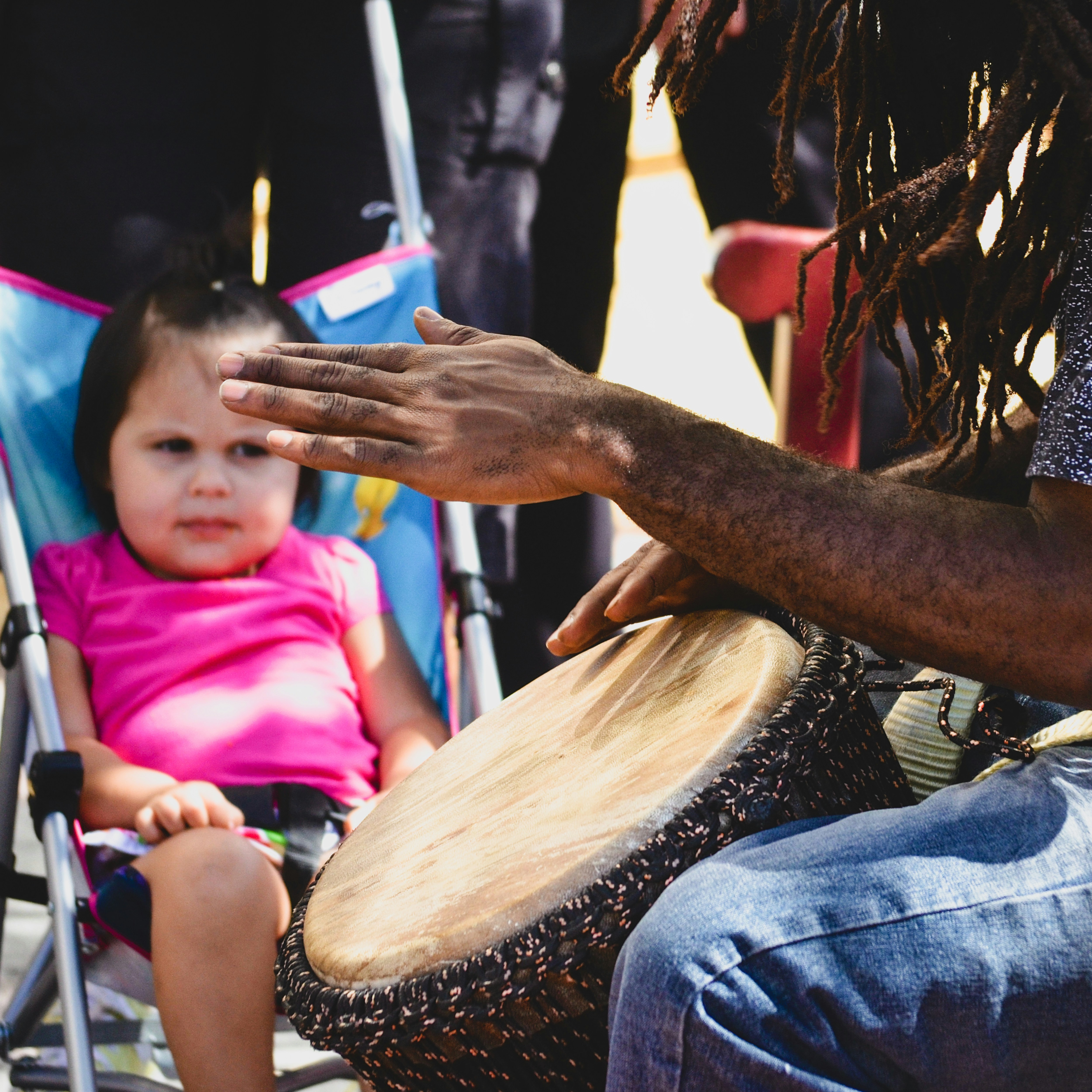 A musician plays a drum while a young girl watches intently, showcasing a moment of shared joy and cultural exchange.