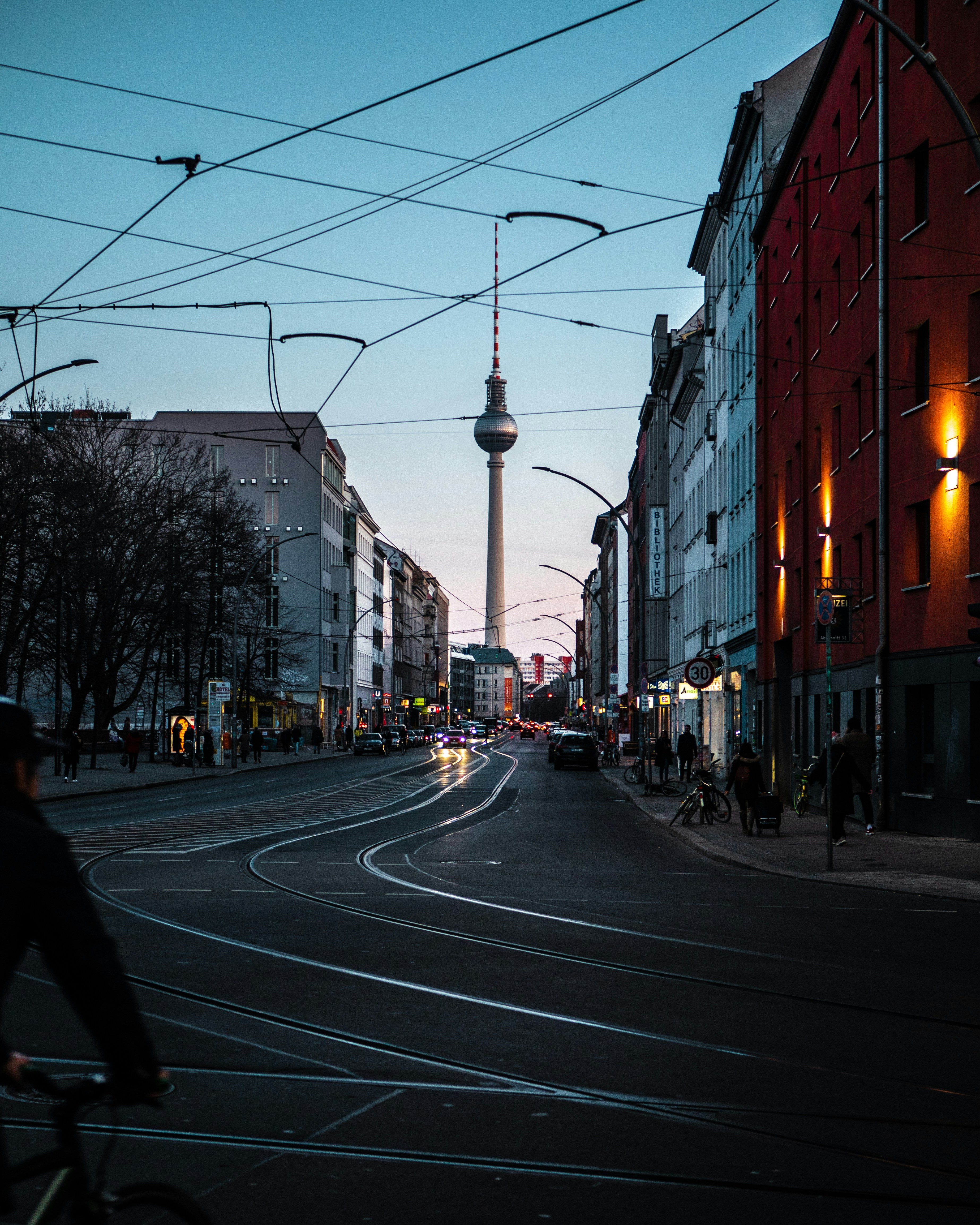 The iconic Berlin TV Tower rises above the cityscape, framed by streetcars and illuminated buildings during twilight.