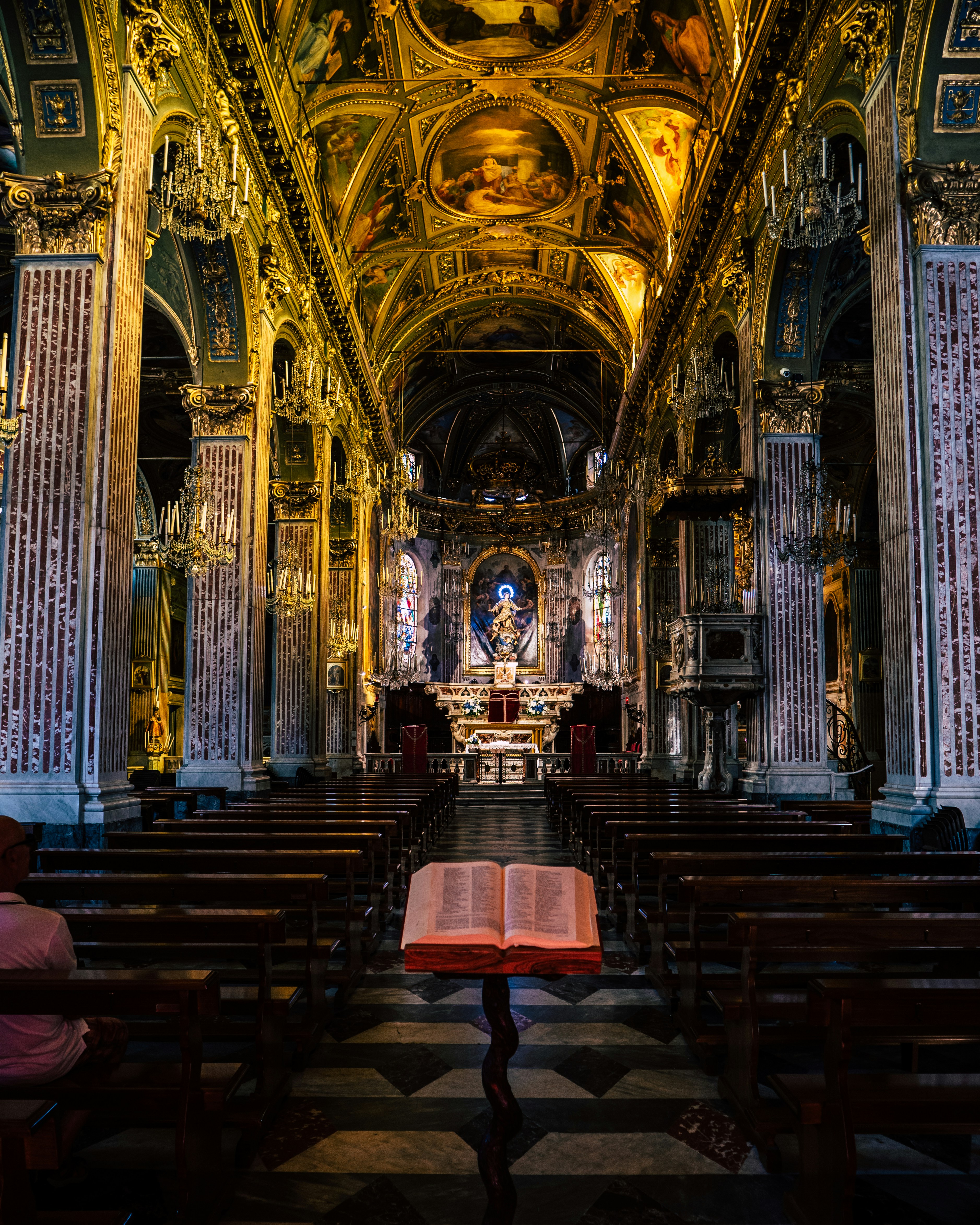 An ornate interior of a church showcasing intricate architecture and a central altar, with an open book in the foreground. The play of light accentuates the details of the surroundings.