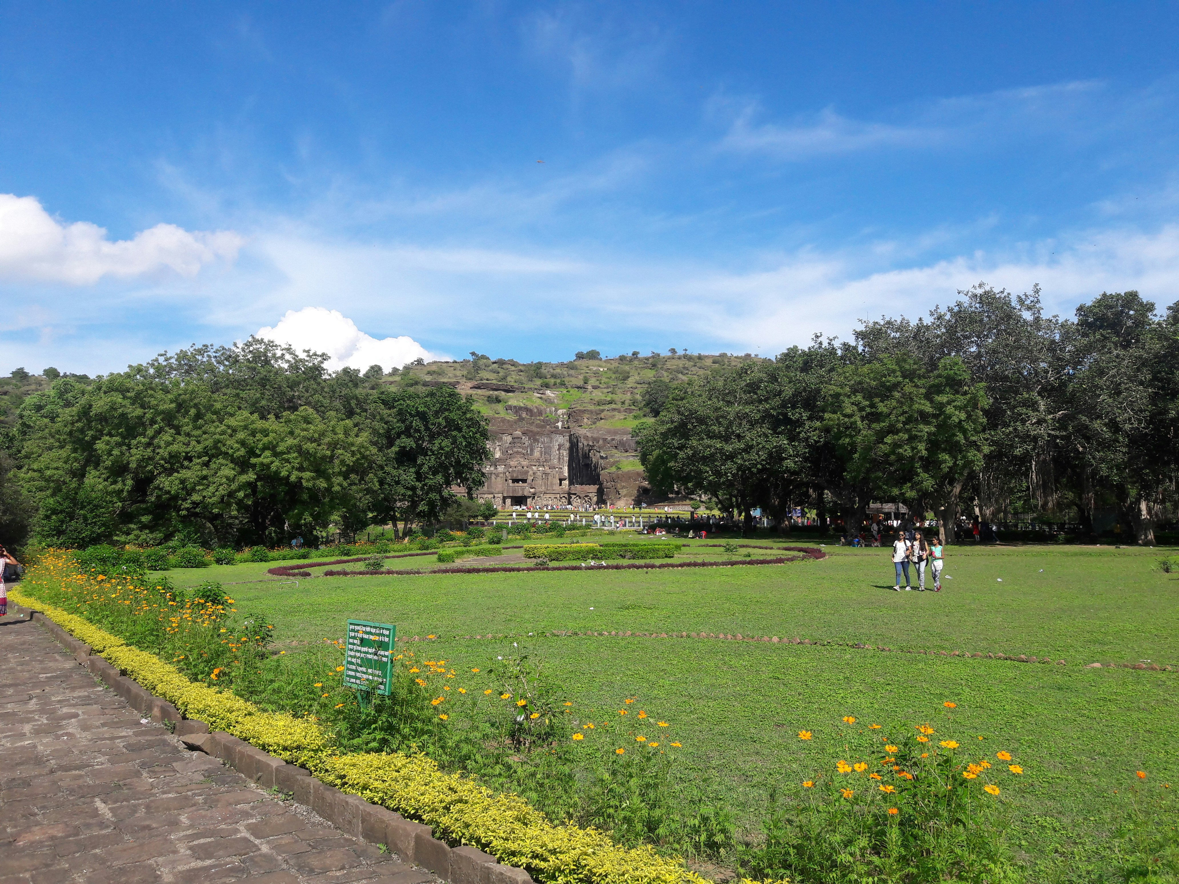 Lush green gardens with vibrant flowers leading toward historic rock-cut caves under a bright blue sky.