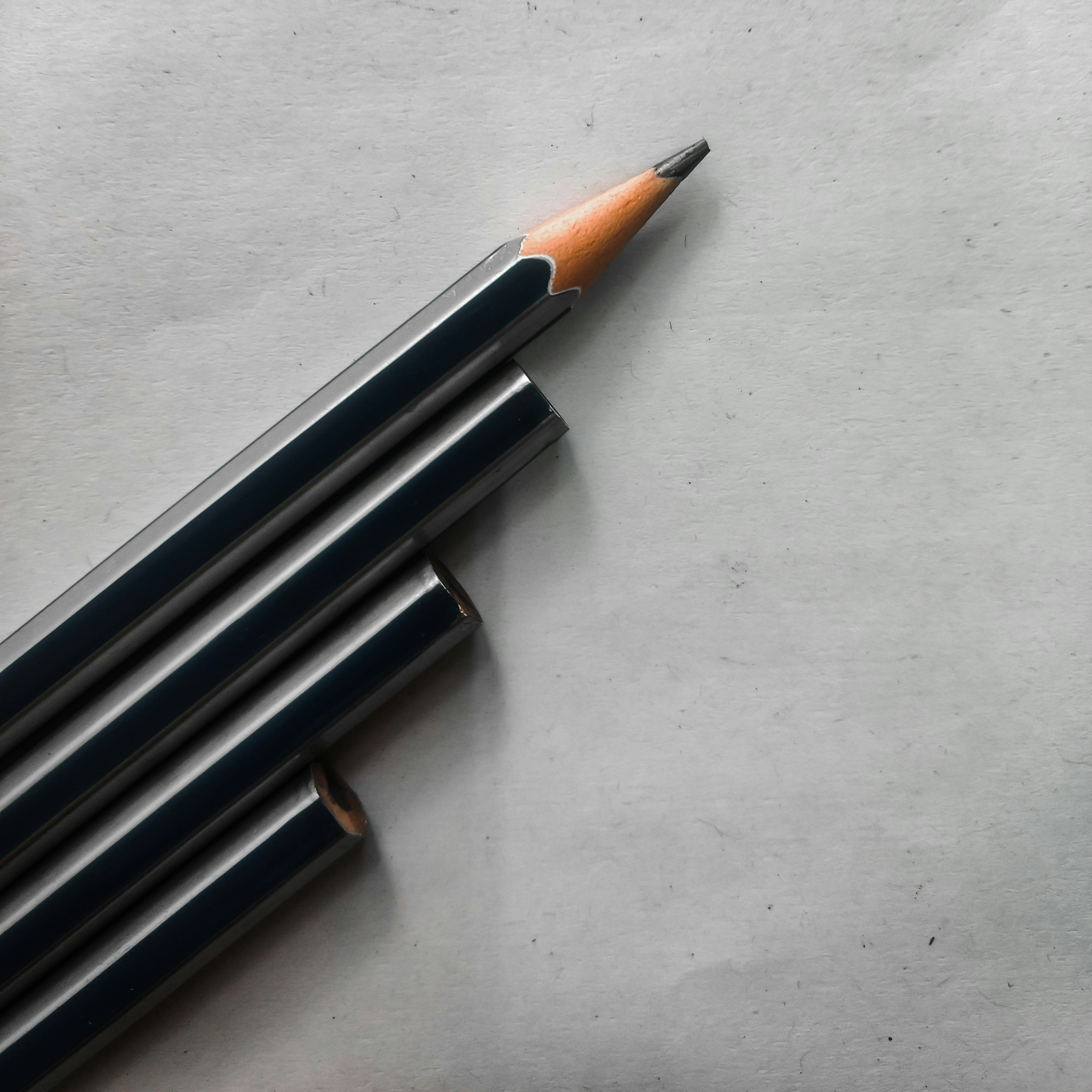 Photograph of three dark pencils with sharpened orange tips arranged diagonally on a textured gray surface. The composition emphasizes color contrast and precise pencil geometry.
