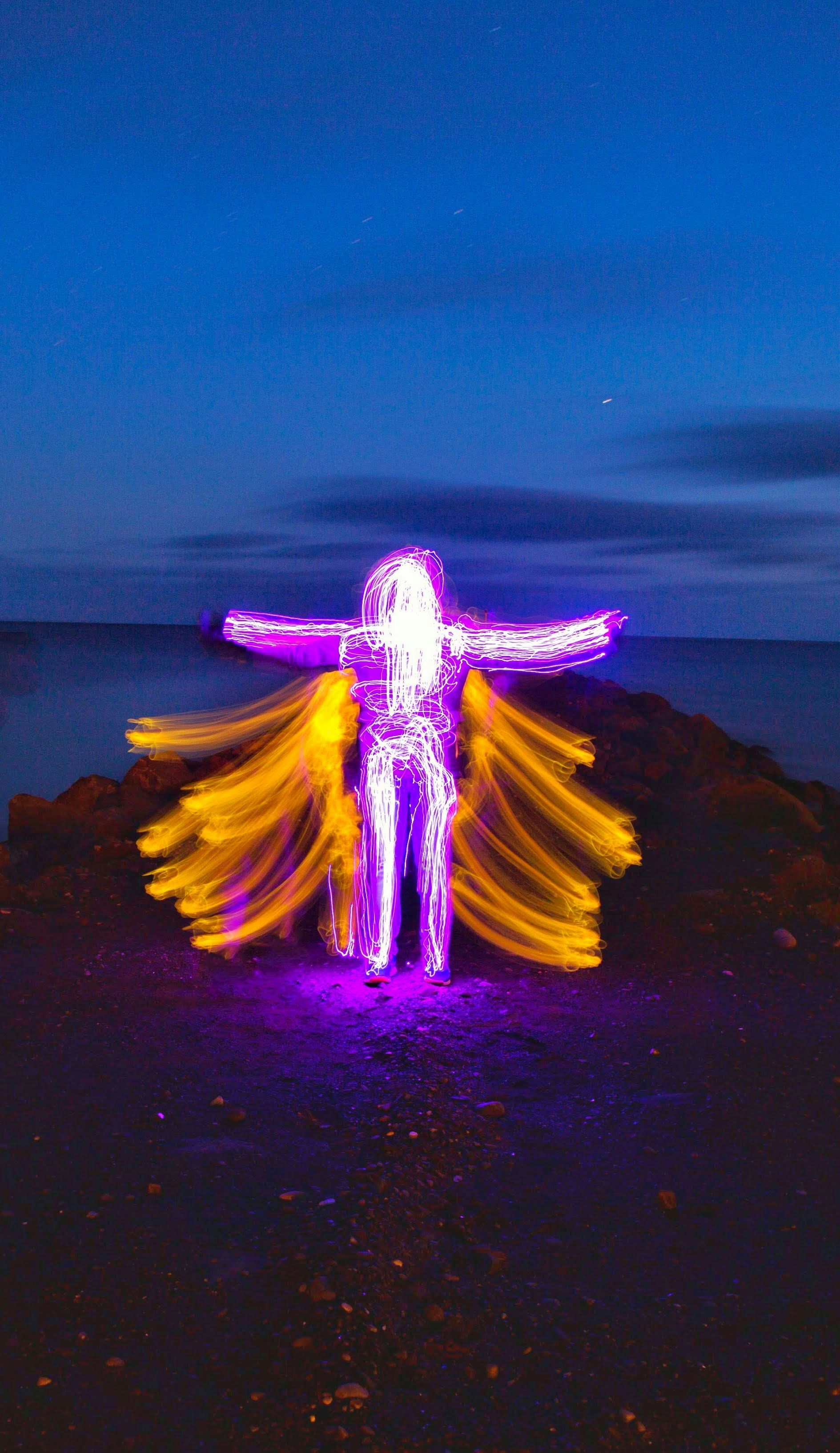 A luminous figure with flowing light trails stands against the twilight sea, embodying movement and energy.