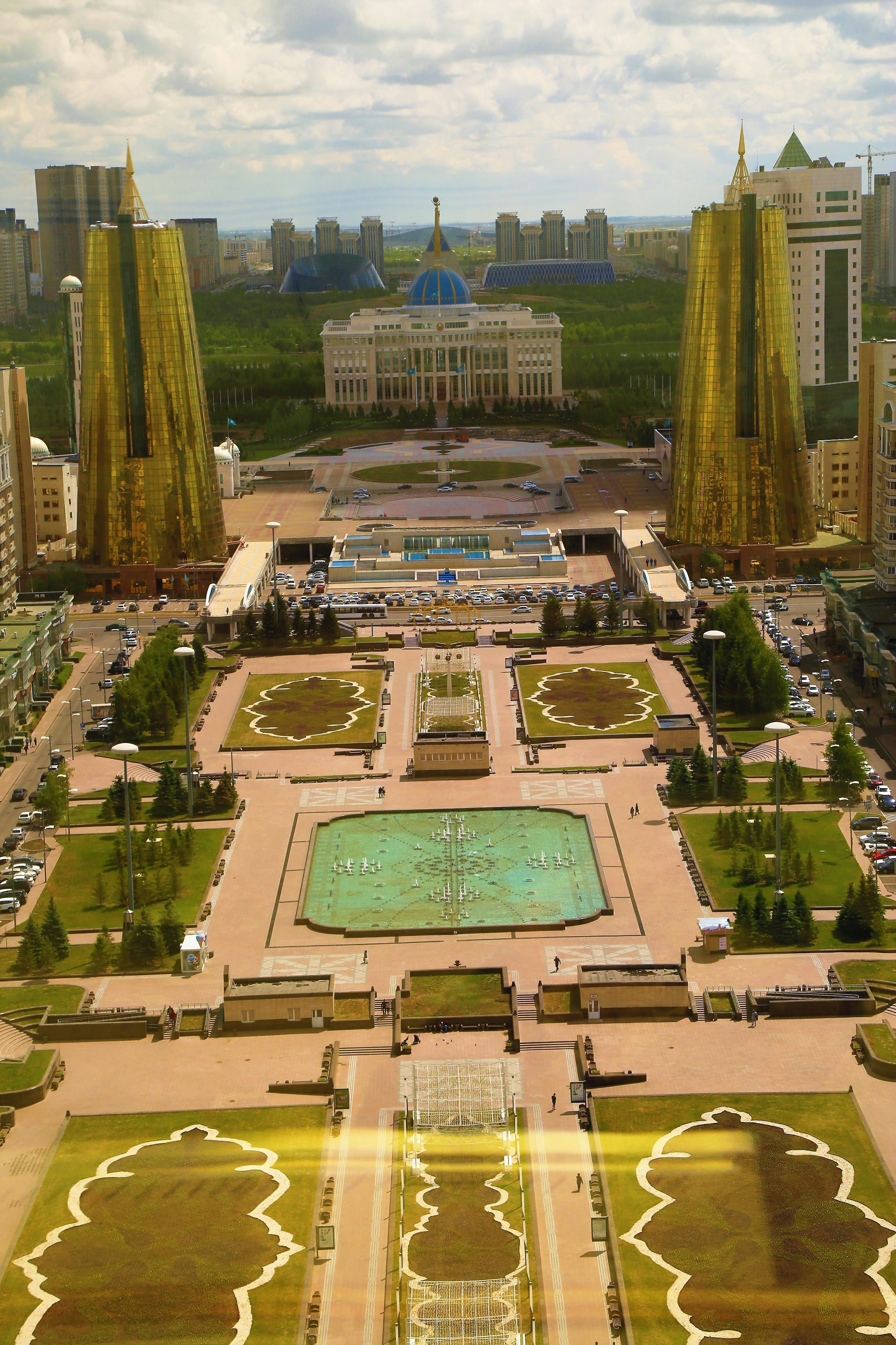 Aerial view of a vibrant city square featuring ornate gardens, a central fountain, and striking golden skyscrapers against a backdrop of modern architecture.