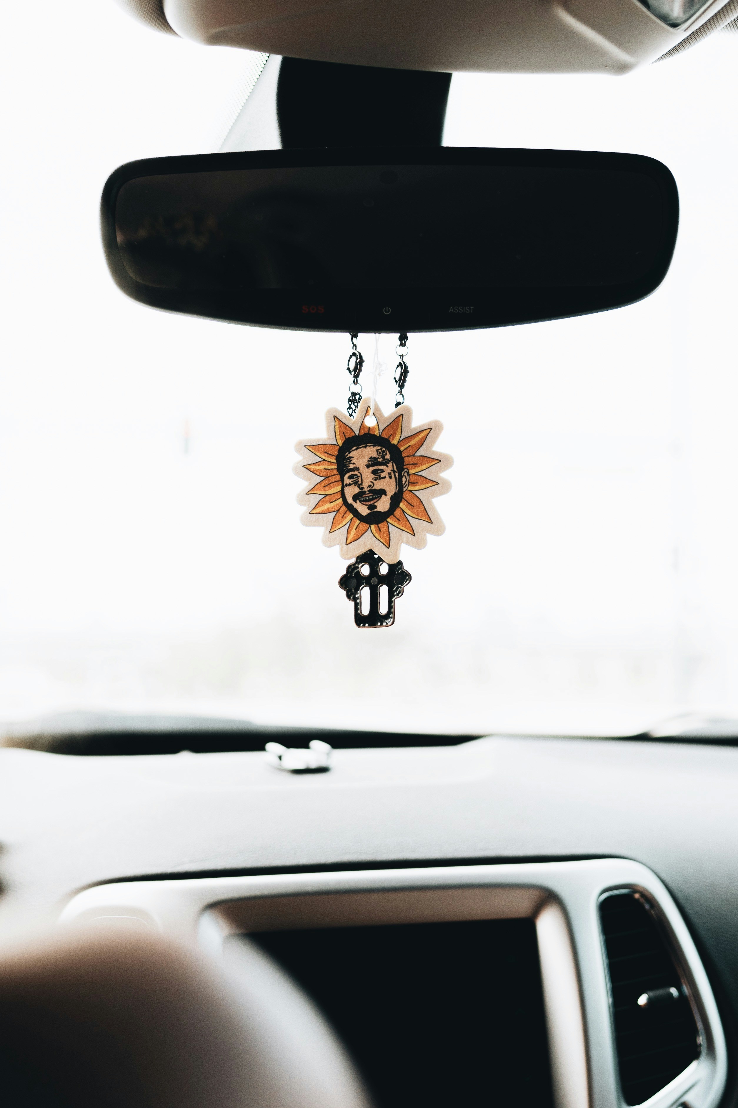 Hanging ornament featuring a sun design with a face, suspended from a car's rearview mirror against a bright background.