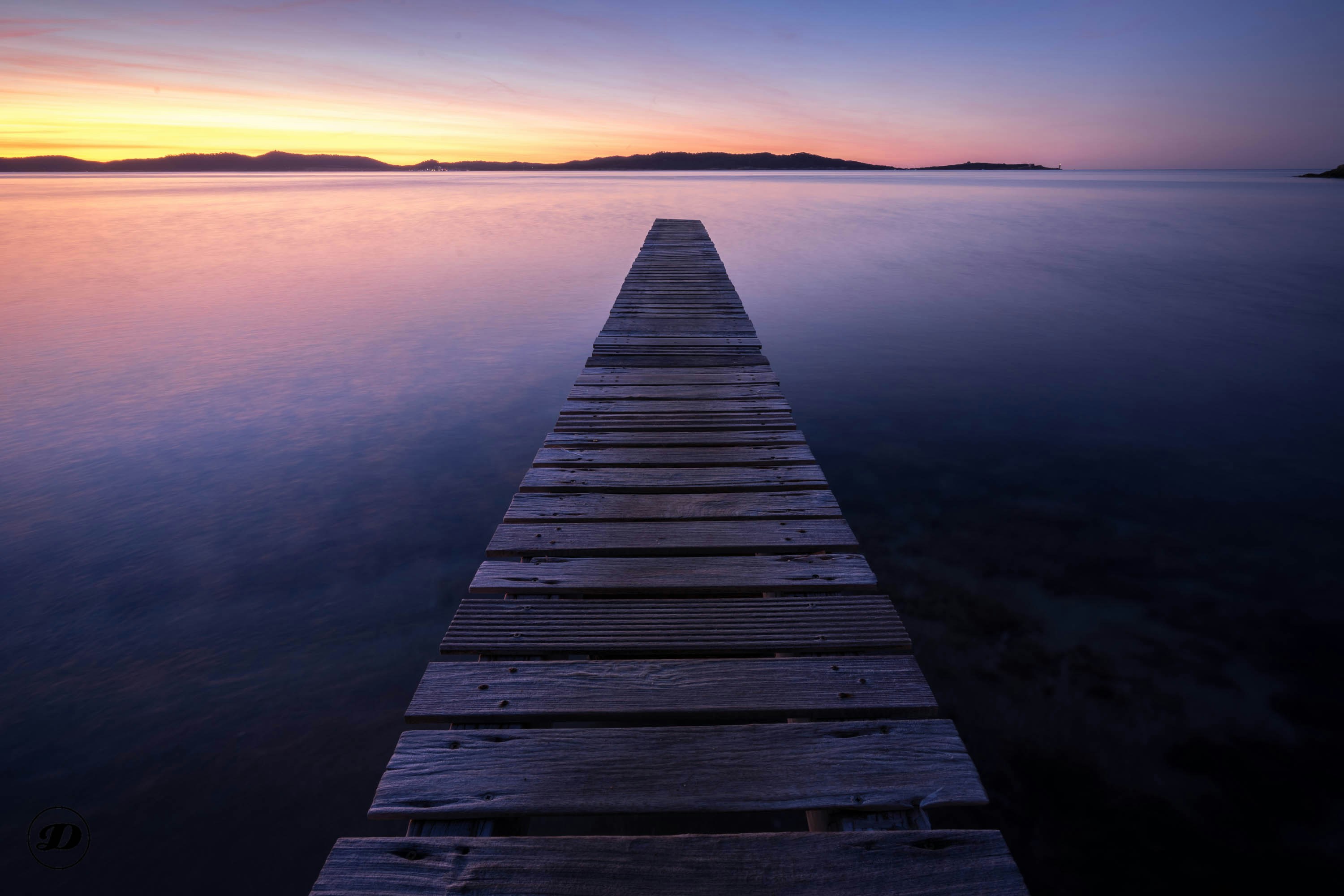 Long wooden dock extending into a tranquil body of water under a vibrant sunset sky.