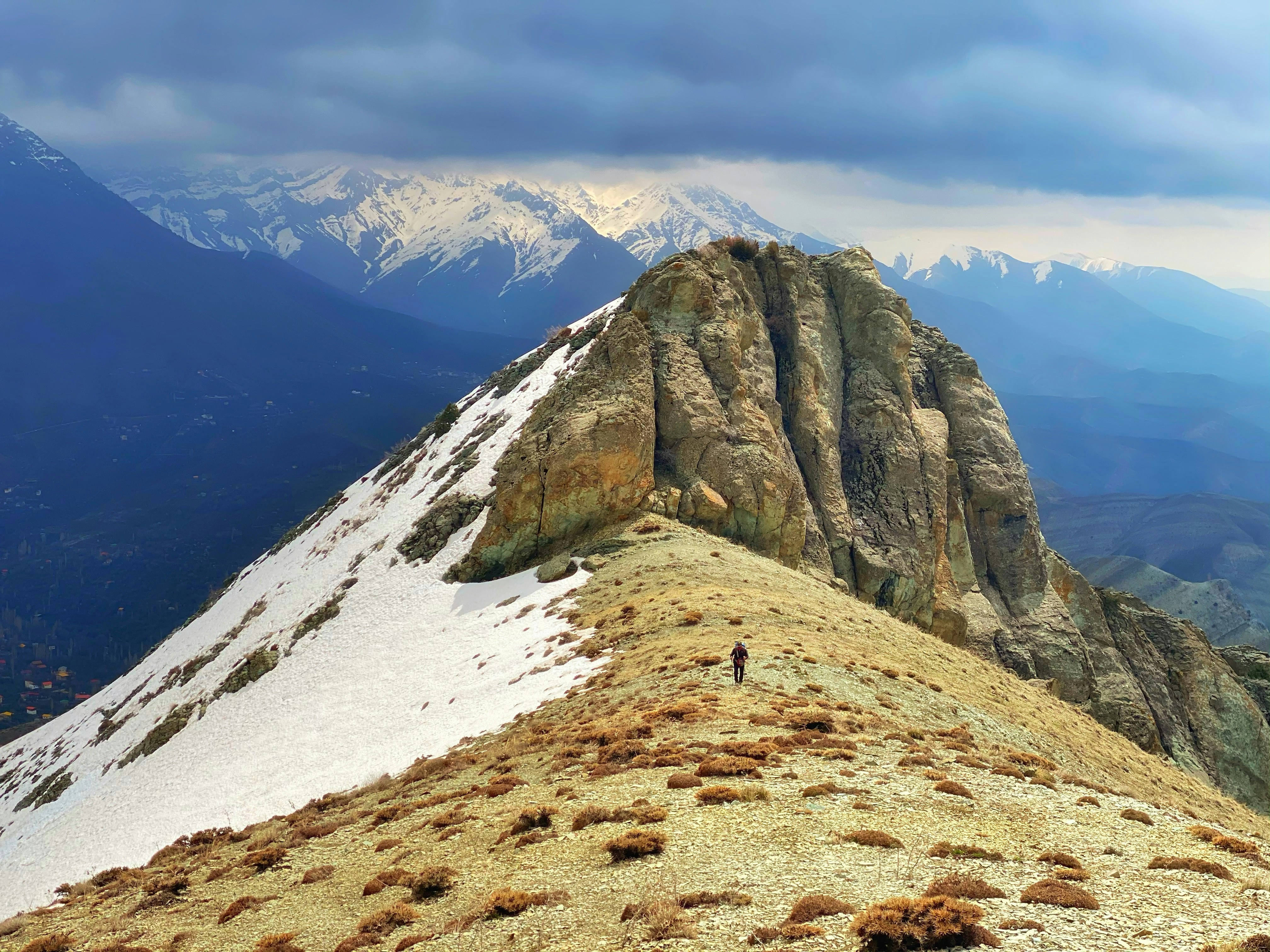 Hiker traversing a rocky ridge with snow patches, surrounded by majestic mountain peaks under a moody sky.