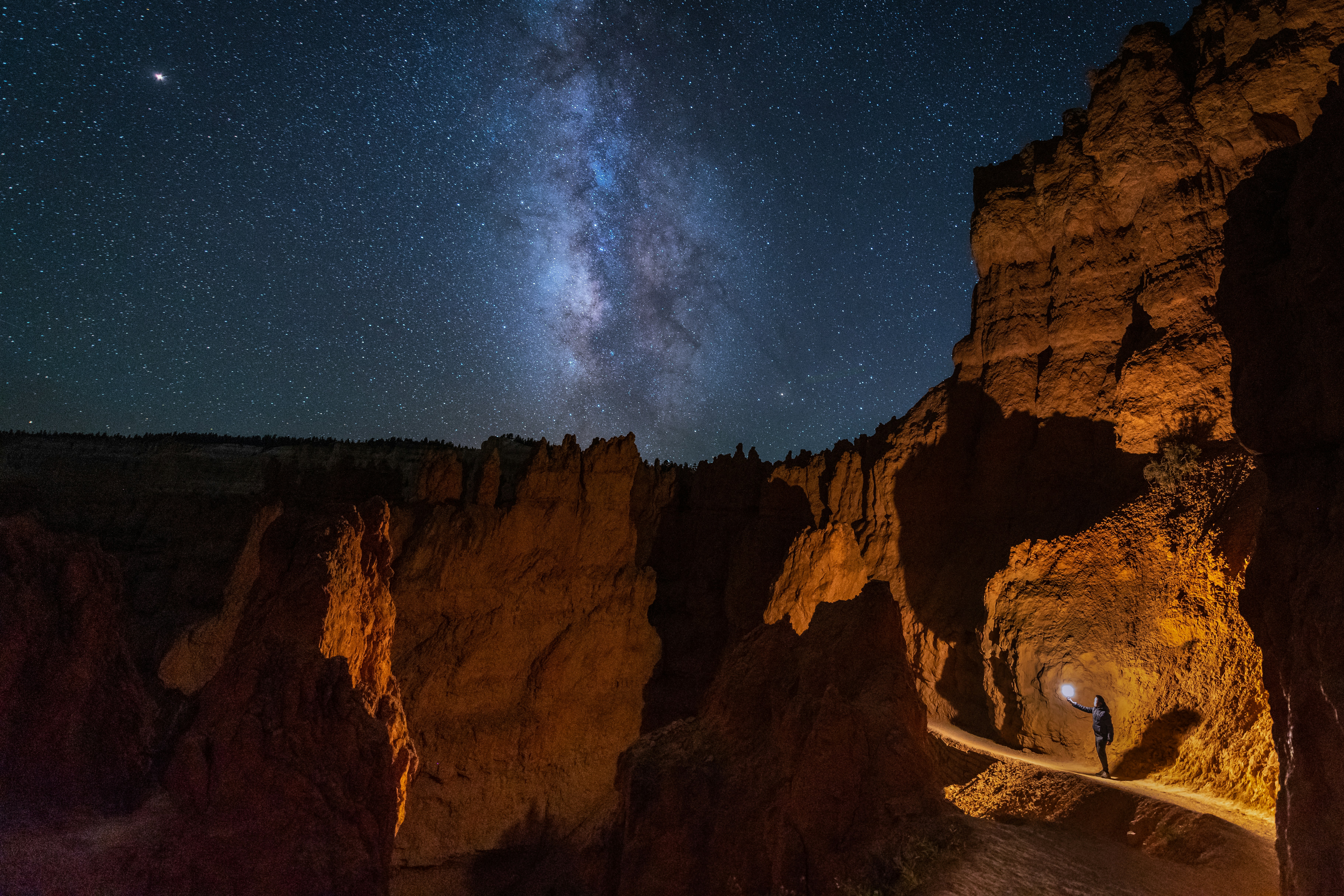 Milky Way galaxy arches over illuminated rock formations and a hiker with a flashlight in a canyon.