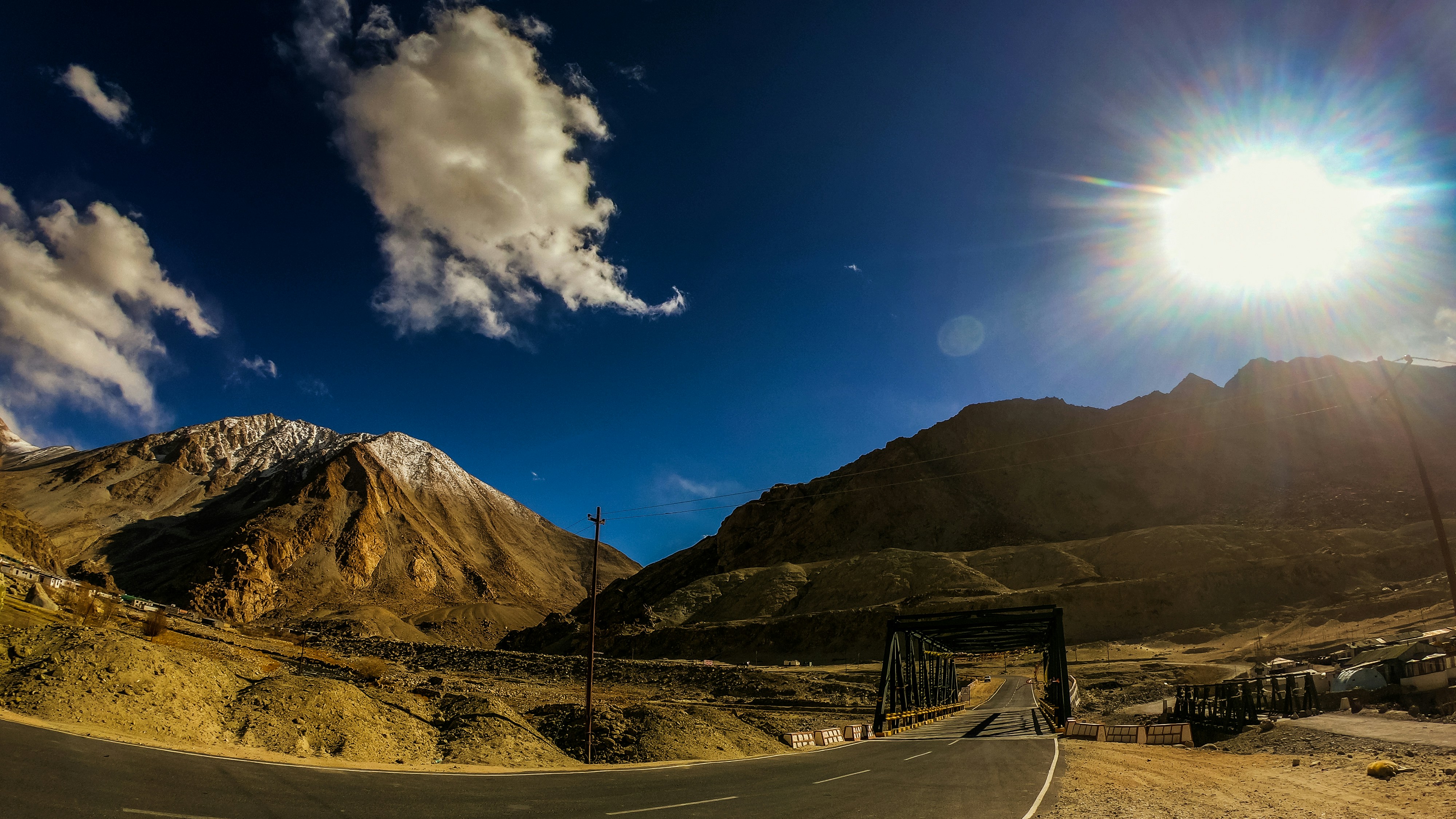 Sunlit mountain road winding through arid landscape with scattered clouds in a vibrant blue sky.