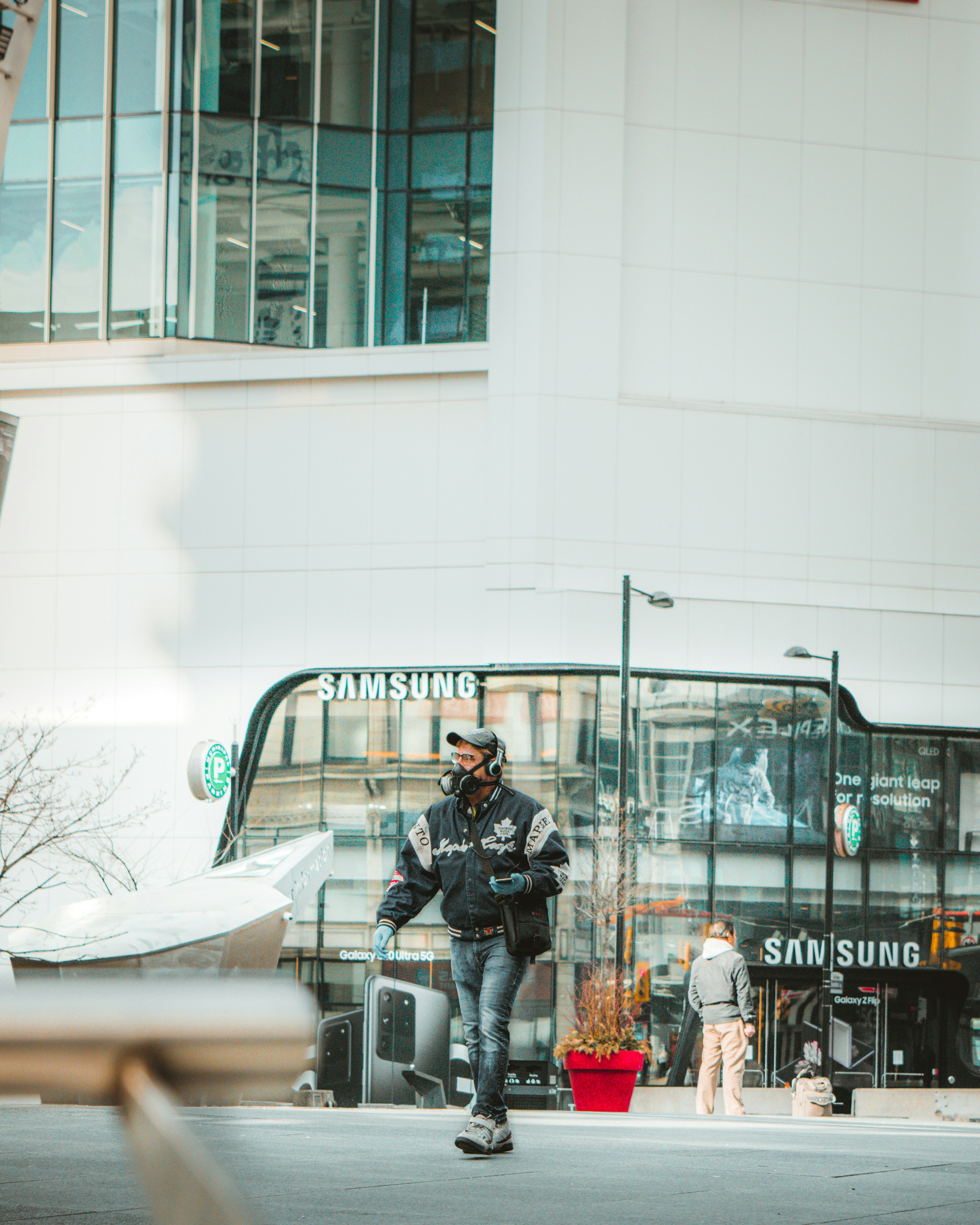 Man in a stylish jacket walking through a bustling urban area with a Samsung storefront in the background. The scene captures the essence of city life.