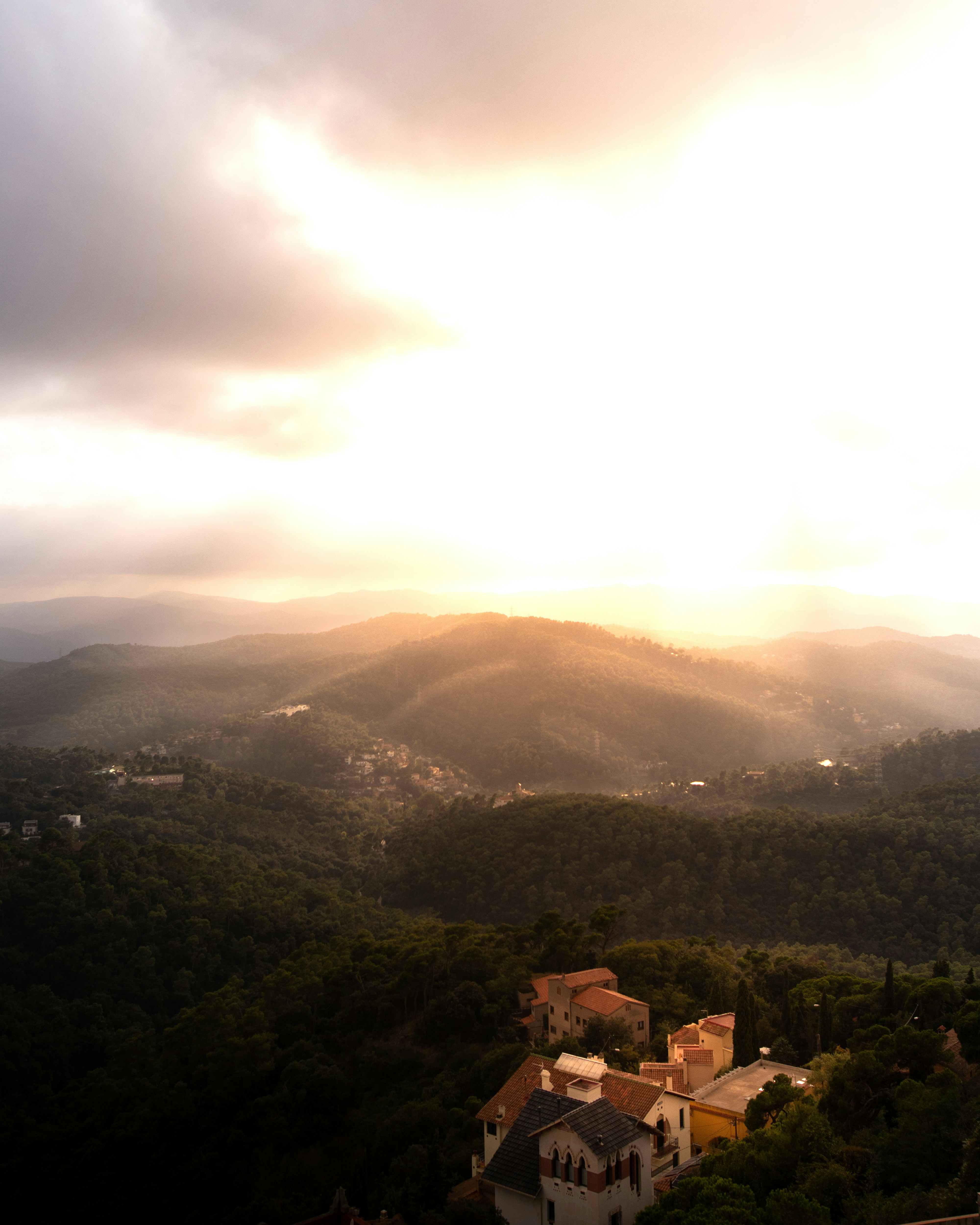 Golden light breaks through clouds, illuminating a serene valley dotted with quaint houses nestled among lush greenery.
