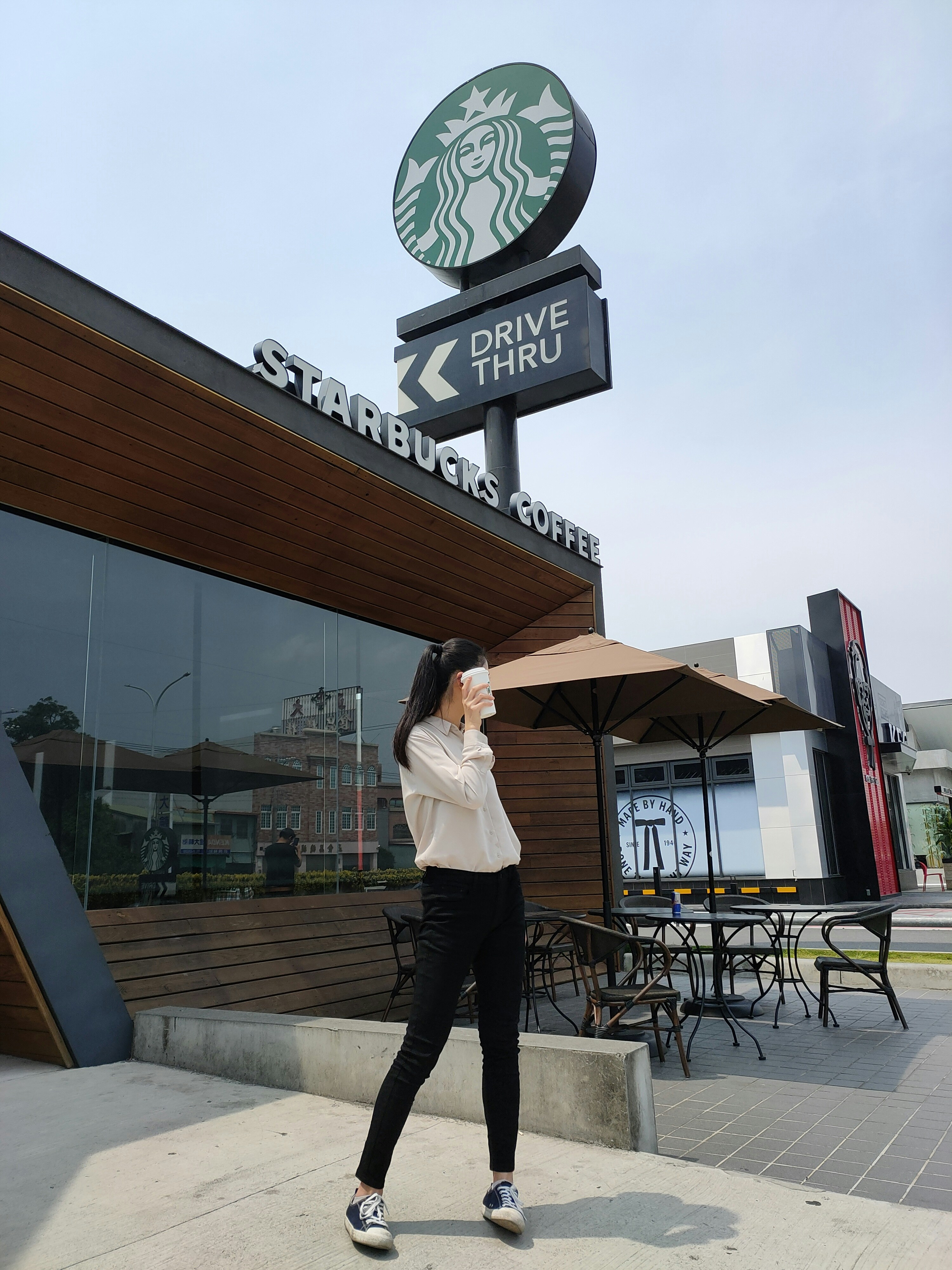 A woman stands outside a modern Starbucks cafe with a drive-thru sign under a clear daytime sky.