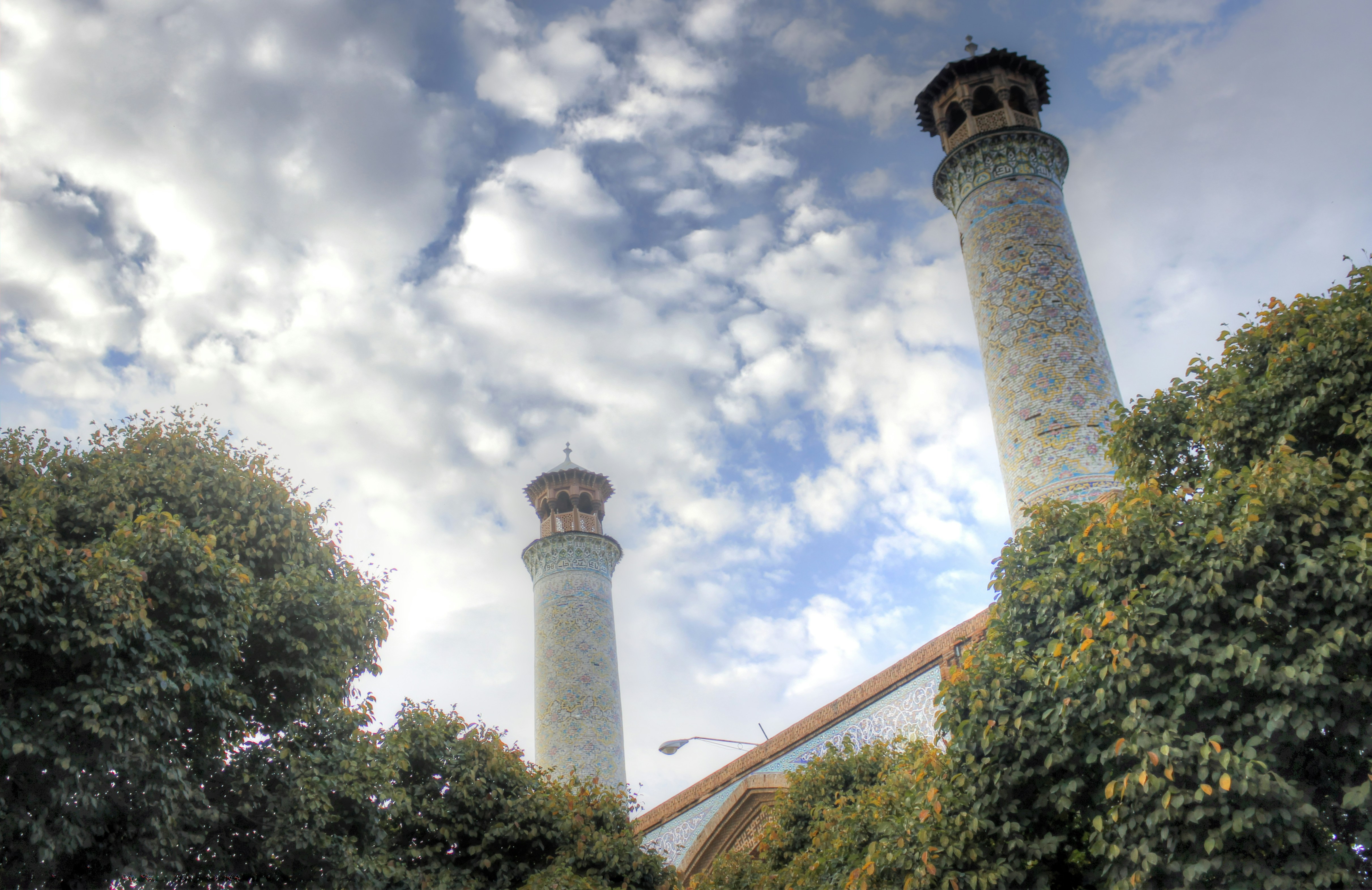 Two ornate towers rise above lush green trees against a partly cloudy sky.