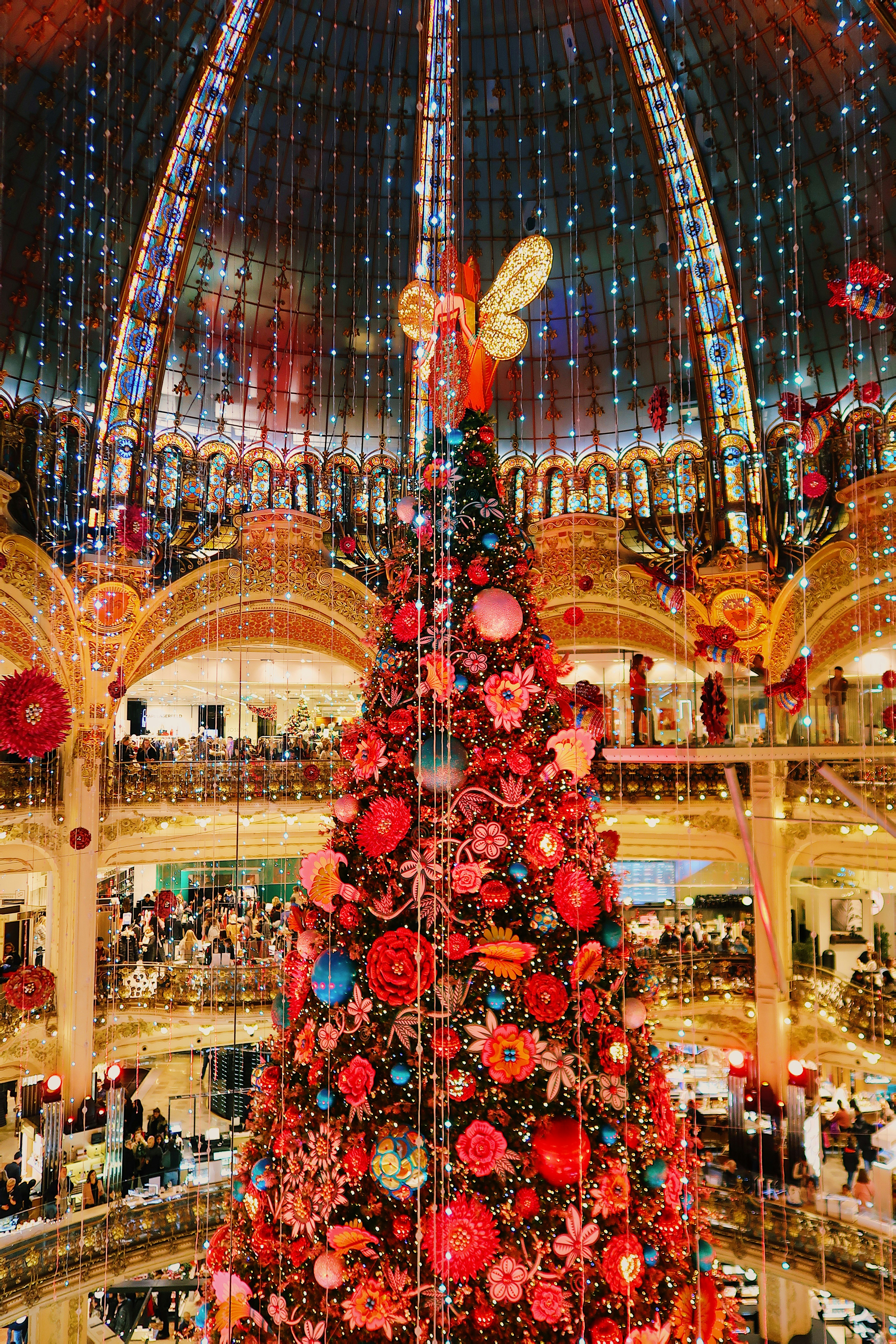 Photograph capturing a grand Christmas tree adorned with vibrant ornaments in an ornate mall atrium. A starry ceiling of hanging lights crowns the scene.