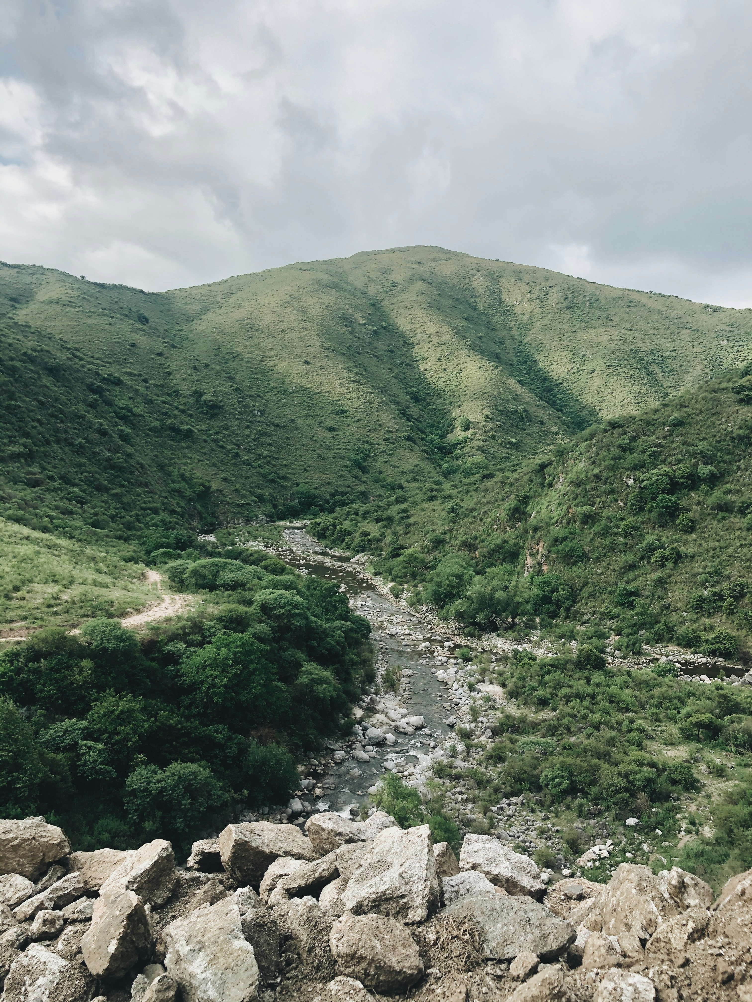 Lush green hills surround a rocky riverbed under a cloudy sky.