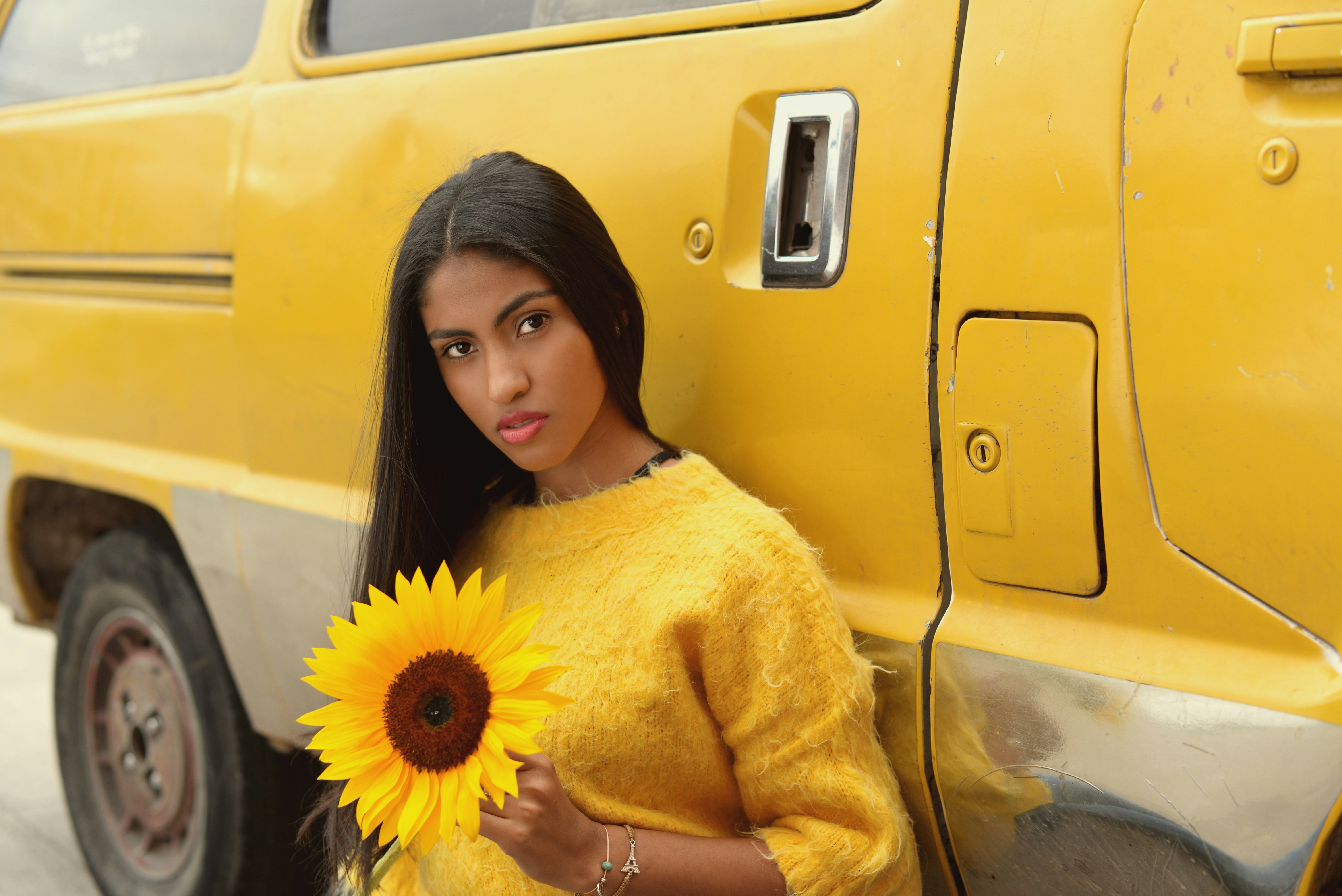 A young woman in a bright yellow sweater holds a sunflower, standing beside a vibrant yellow vehicle, creating a harmonious color palette.