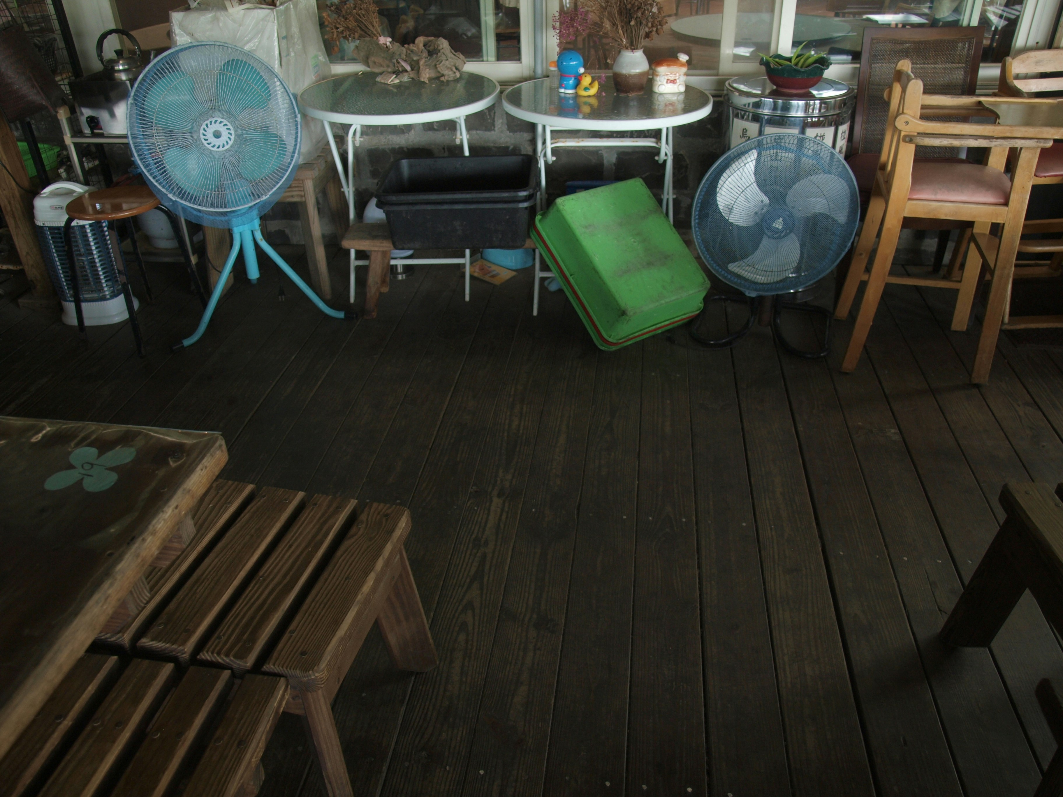 A cozy indoor space featuring various furniture pieces, including fans and tables, arranged in a casual setting. The wooden floor adds warmth to the ambiance.