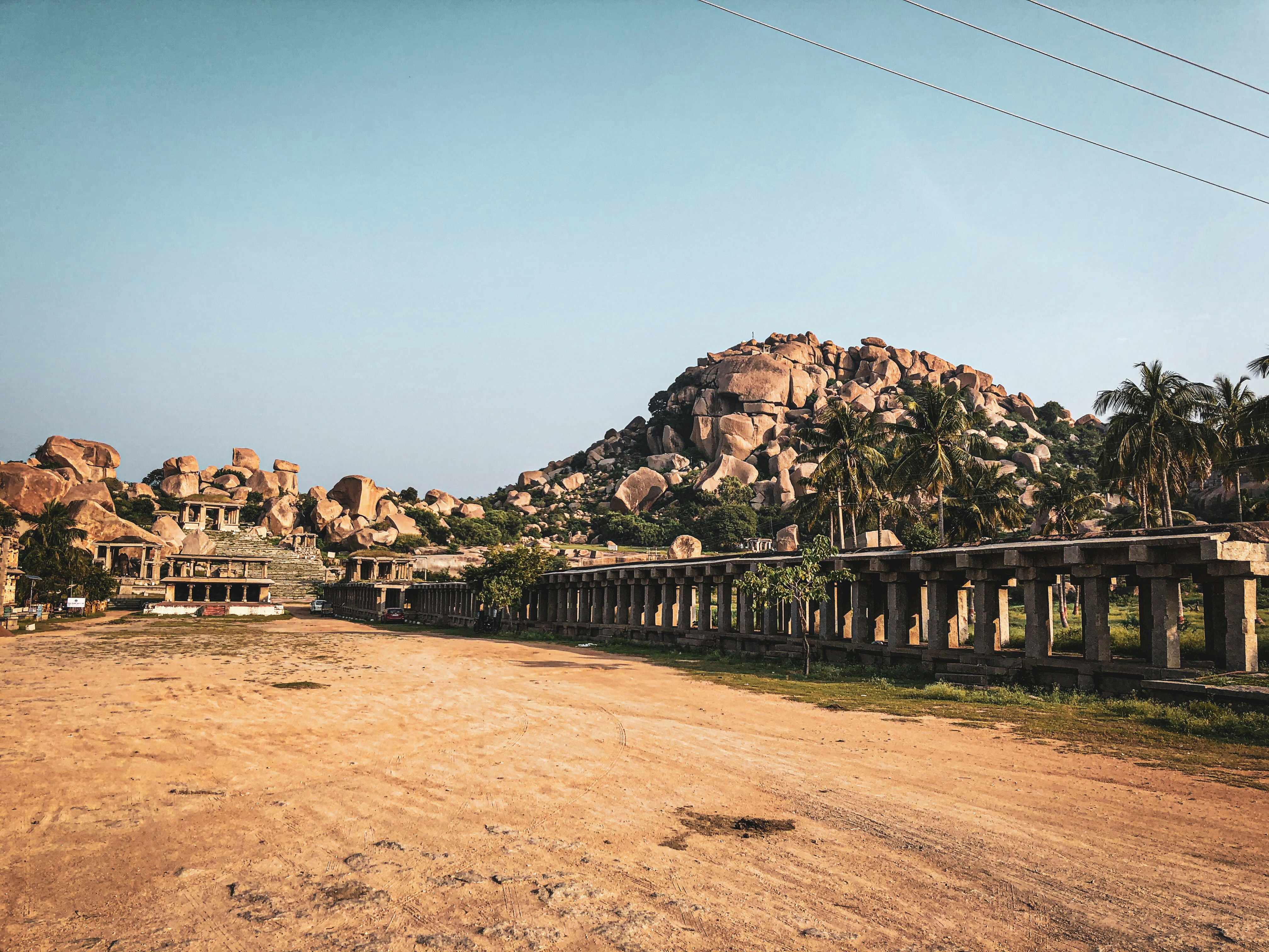 Ruins of a historical structure juxtaposed against a rocky landscape, framed by palm trees under a clear blue sky.
